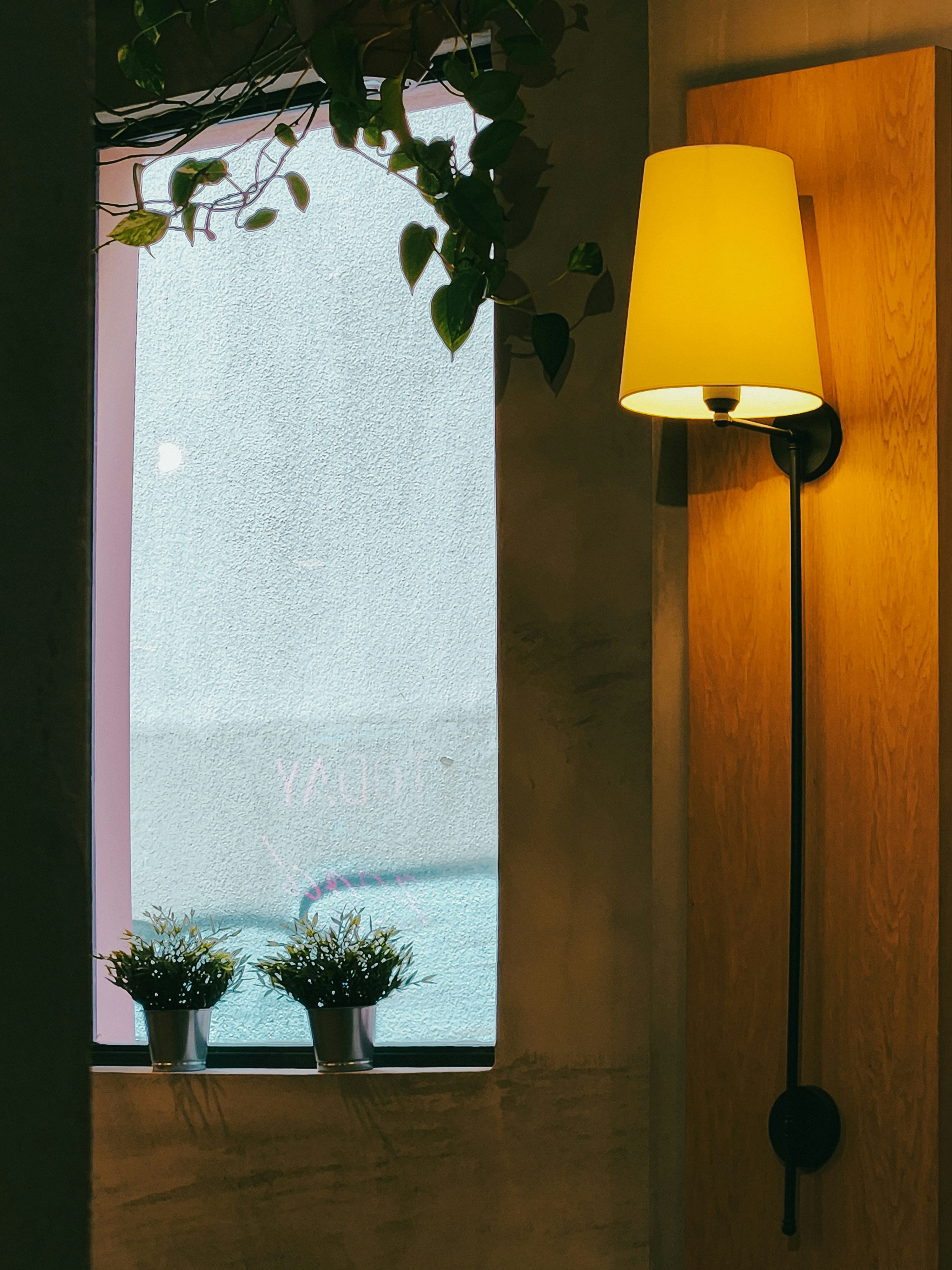 A warm yellow lamp casts a soft glow beside a window adorned with small potted plants, creating a tranquil indoor atmosphere.