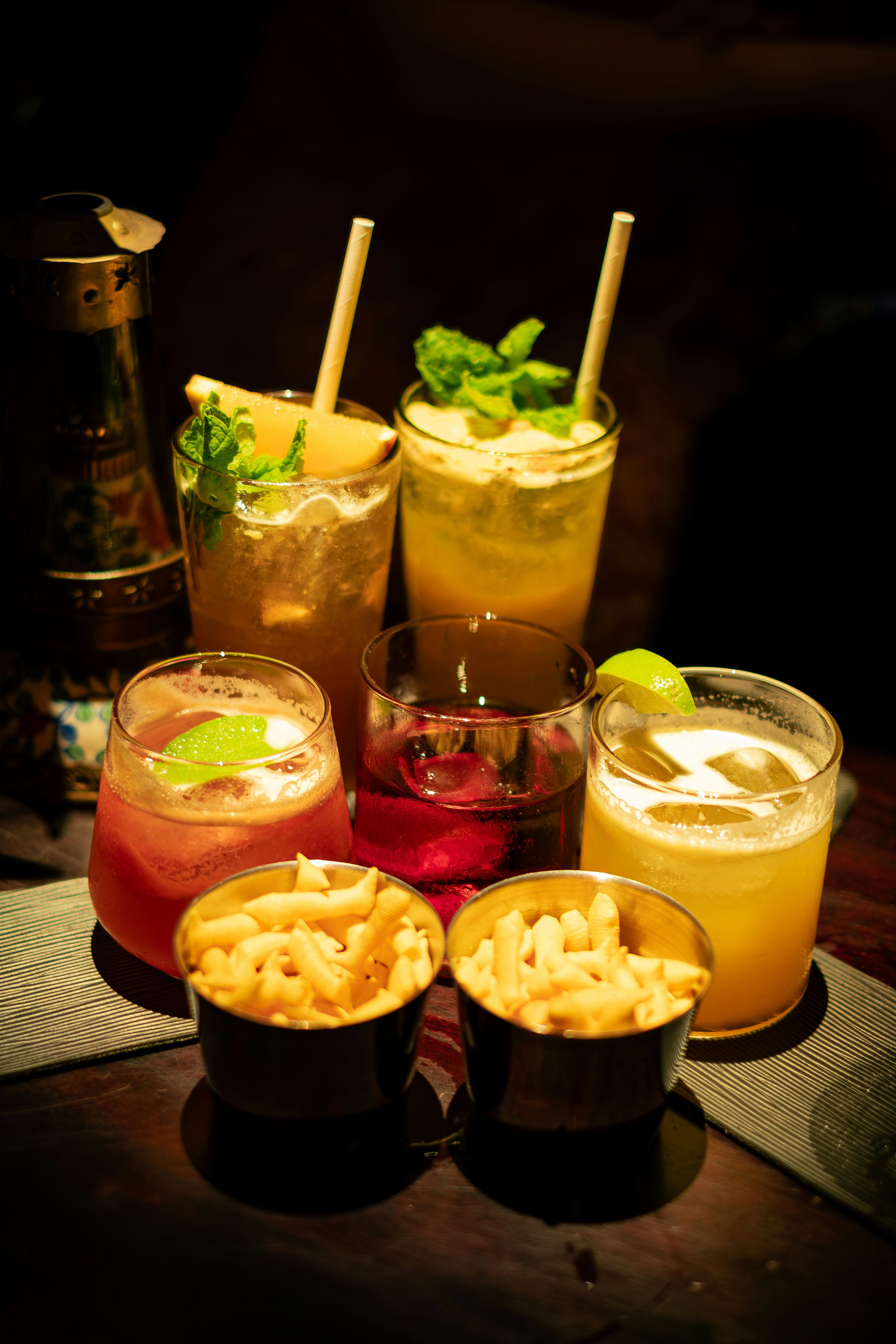 An array of colorful cocktails garnished with fresh herbs, accompanied by bowls of crispy snacks on a wooden table.