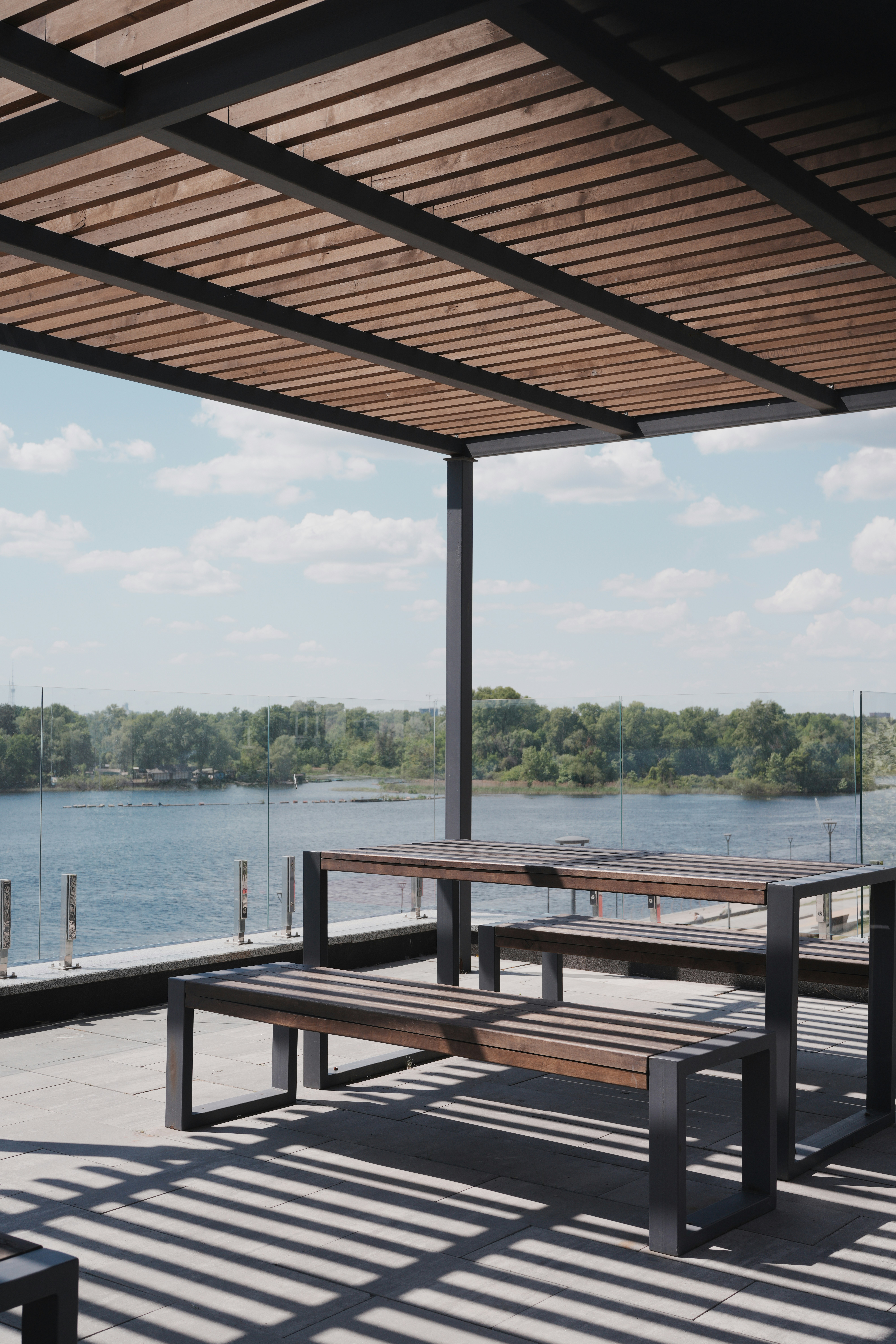 A modern outdoor terrace with wooden benches under a slatted roof overlooks a calm lake; glass railing and shadows create a geometric pattern.