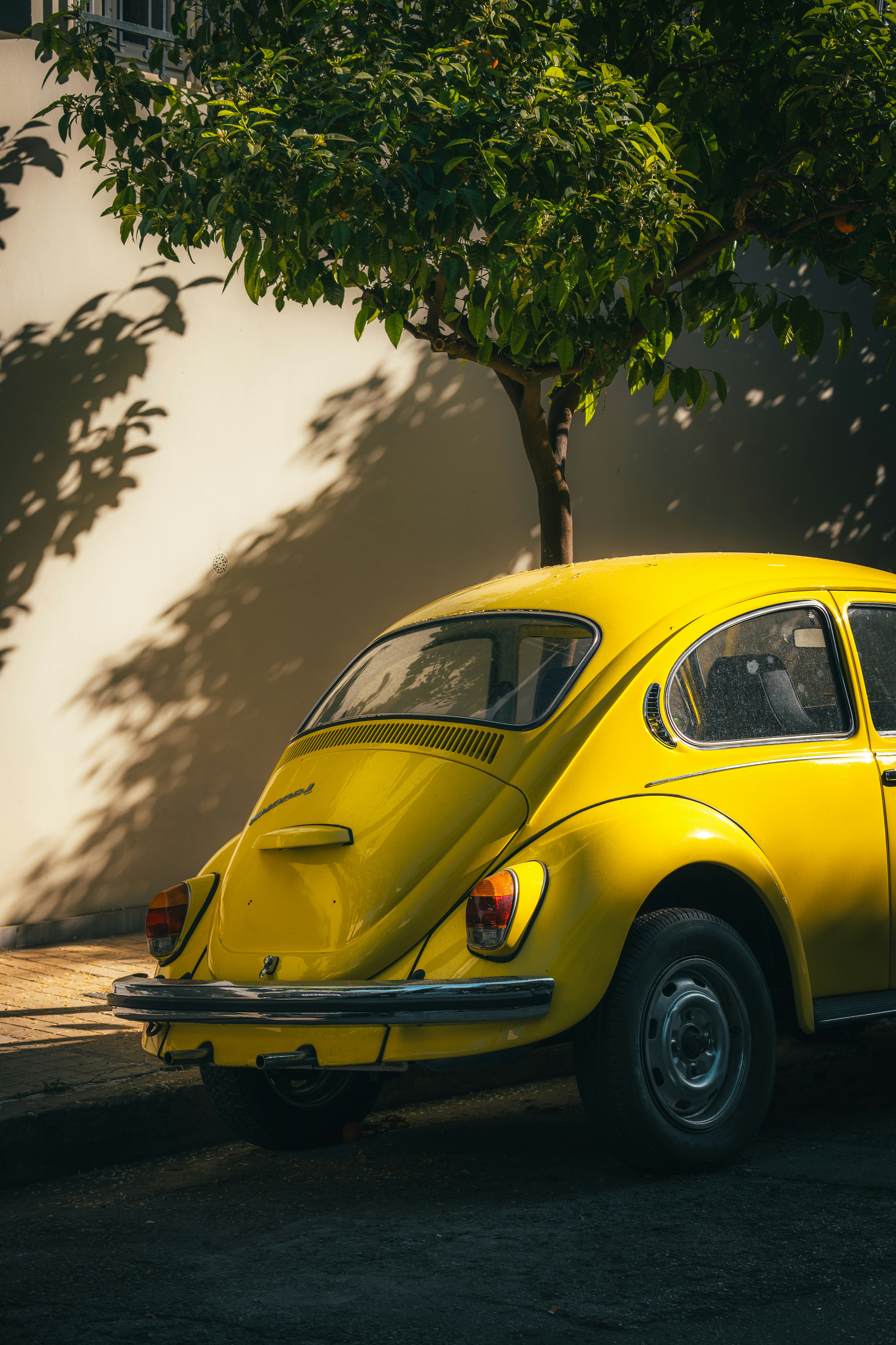 Sunlit yellow vintage car parked beside a shadowed wall beneath a leafy tree.