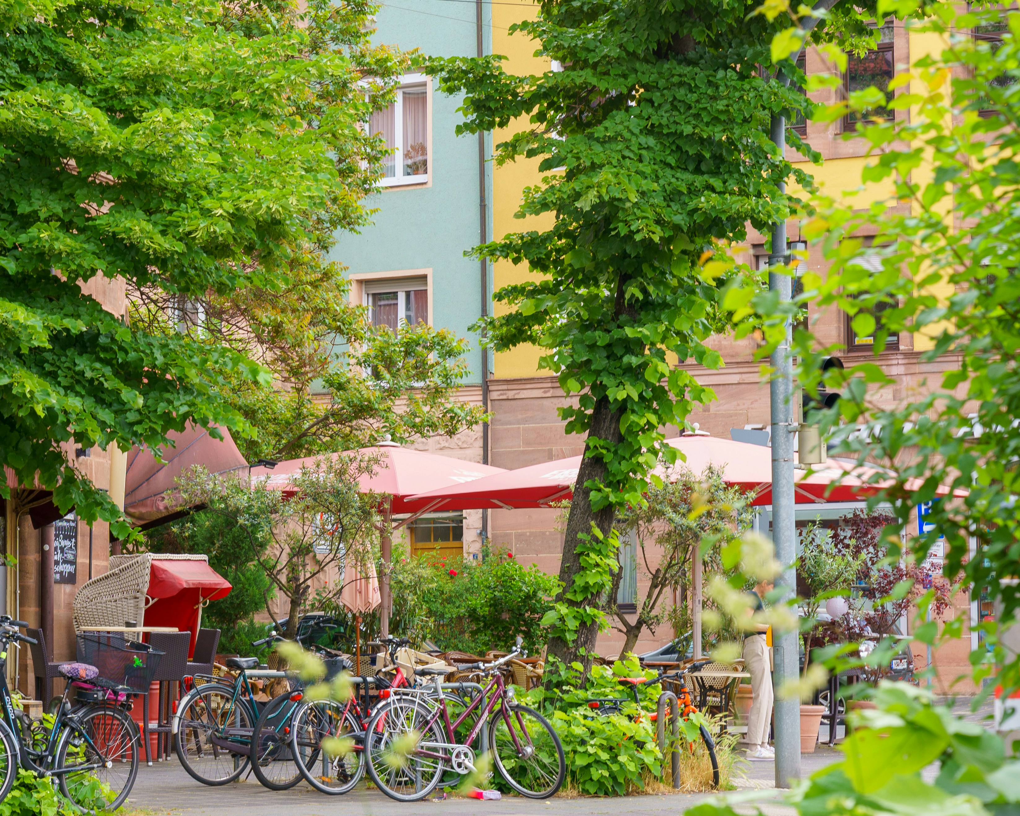Bicycles parked outside a café with red umbrellas, surrounded by lush trees and plants on a sunny day.
