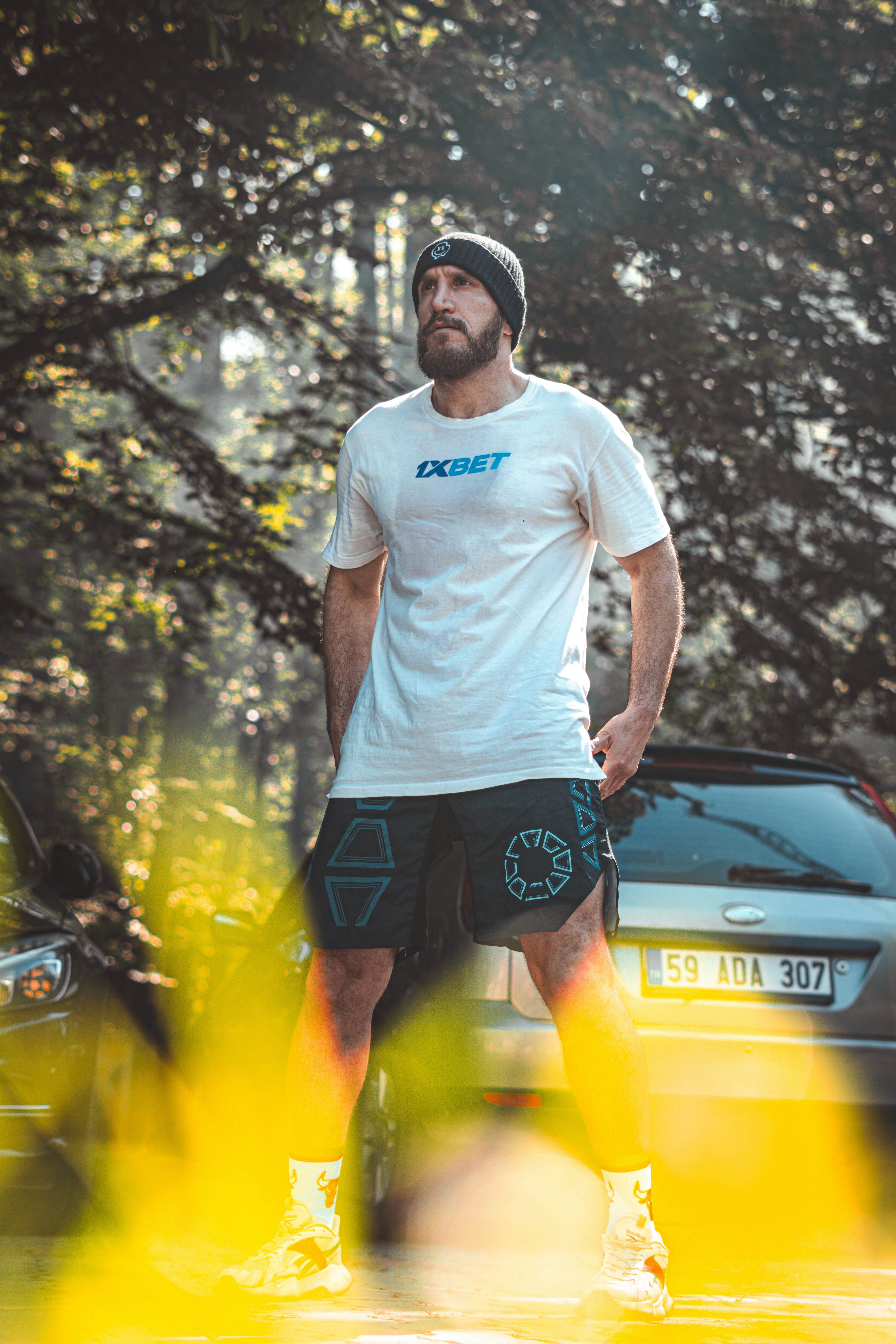 Sunlit Stance: A Bearded Athlete in a White 1XBET Tee Stands Amid Golden Foreground Bokeh