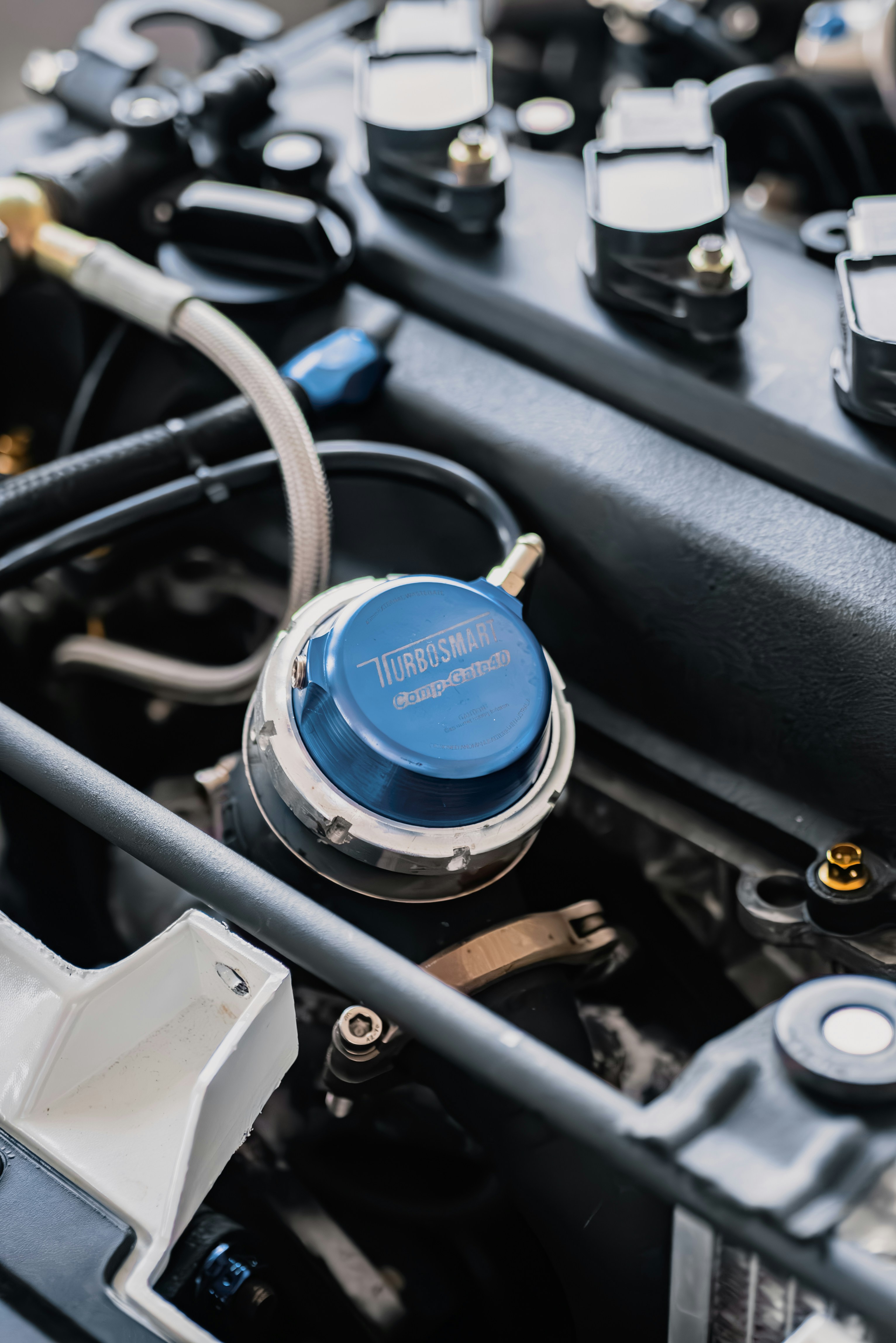 Close-up of a blue TurboSmart cap nestled among engine components, hoses, and metal fittings in a car's engine bay; emphasizes mechanical detail and branding. (Logo appears on cap.)