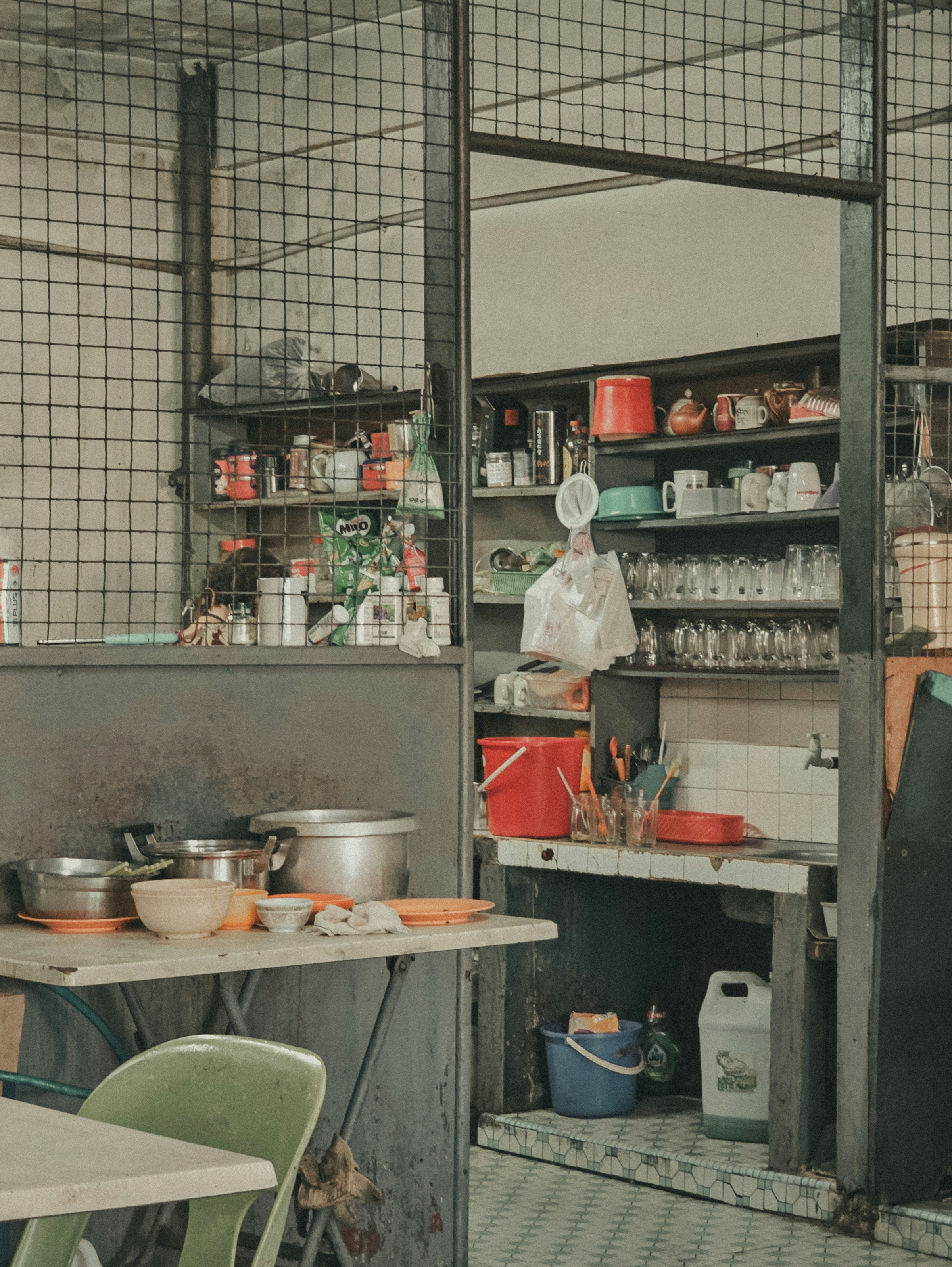 Interior of a cluttered kitchen with metal shelving, stacked dishes, and various containers on a checkered tile floor. A plastic jug and red buckets sit near the doorway, underscoring a lived-in, utilitarian space.