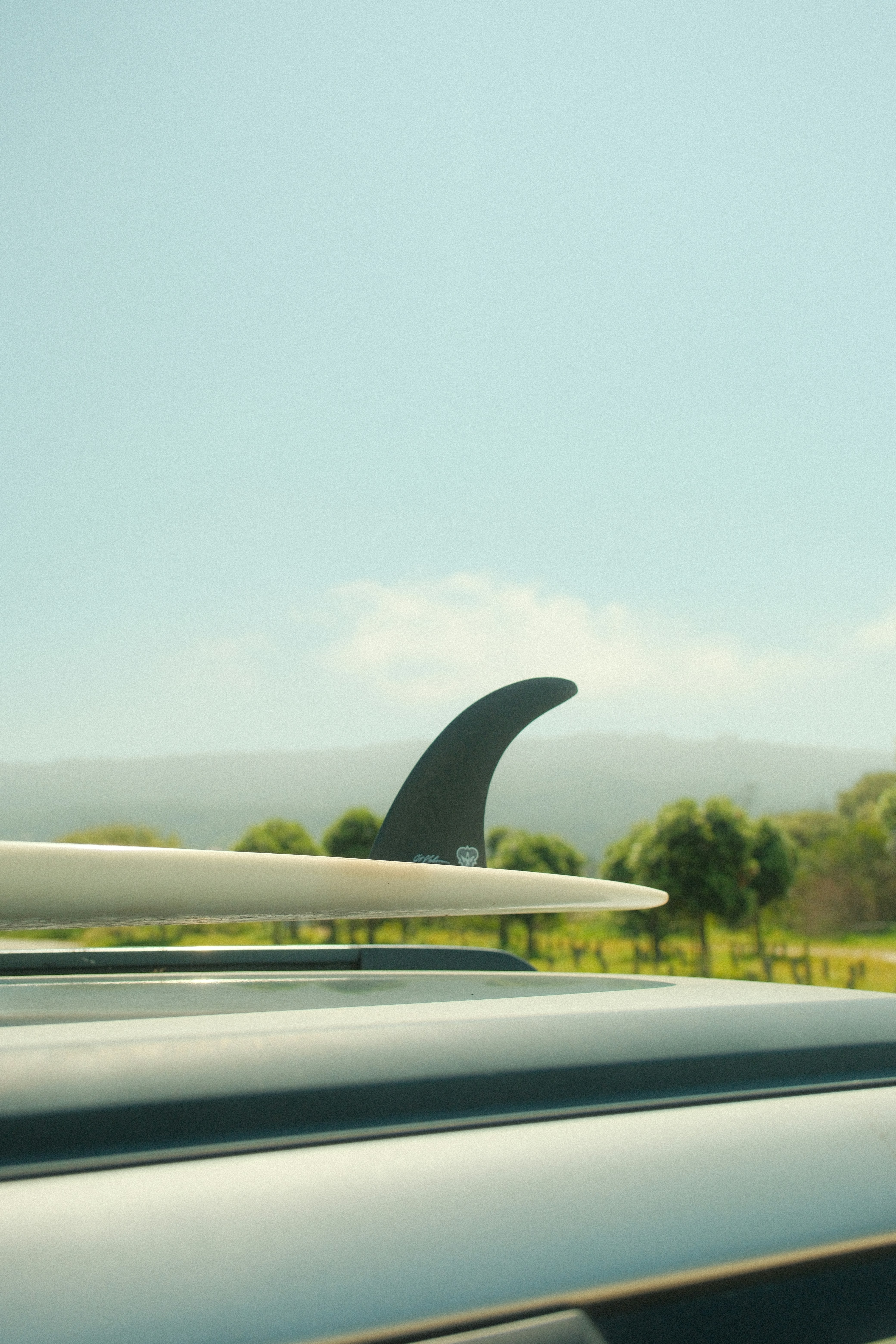 A surfboard with a prominent fin rests atop a vehicle, set against a backdrop of rolling hills and a clear sky. The scene evokes a sense of adventure and readiness for the waves.
