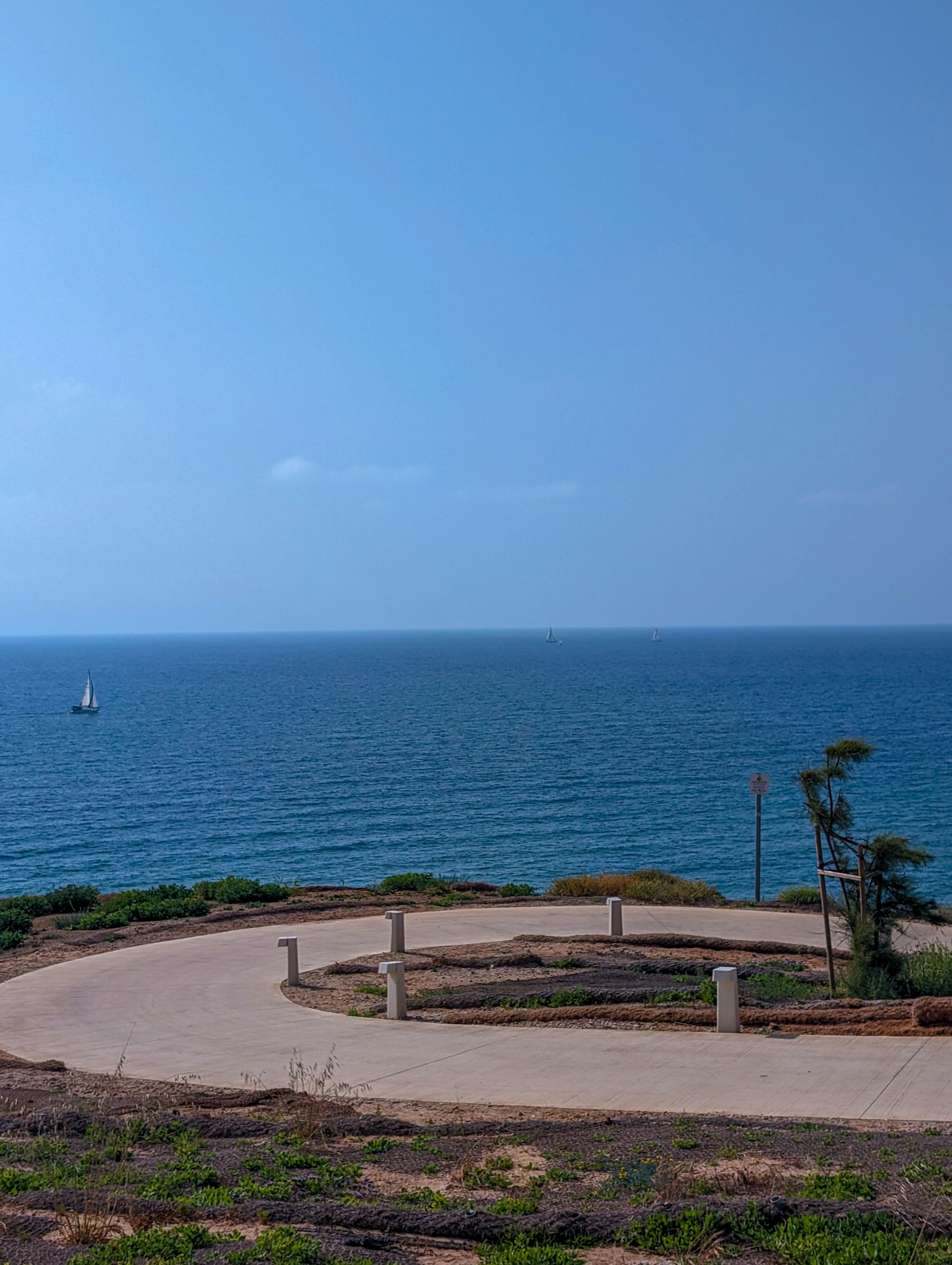 A curved coastal promenade frames the foreground, guiding the eye toward a calm sea and clear blue sky. A small sailboat drifts on the left, adding a gentle focal point to the tranquil seascape.