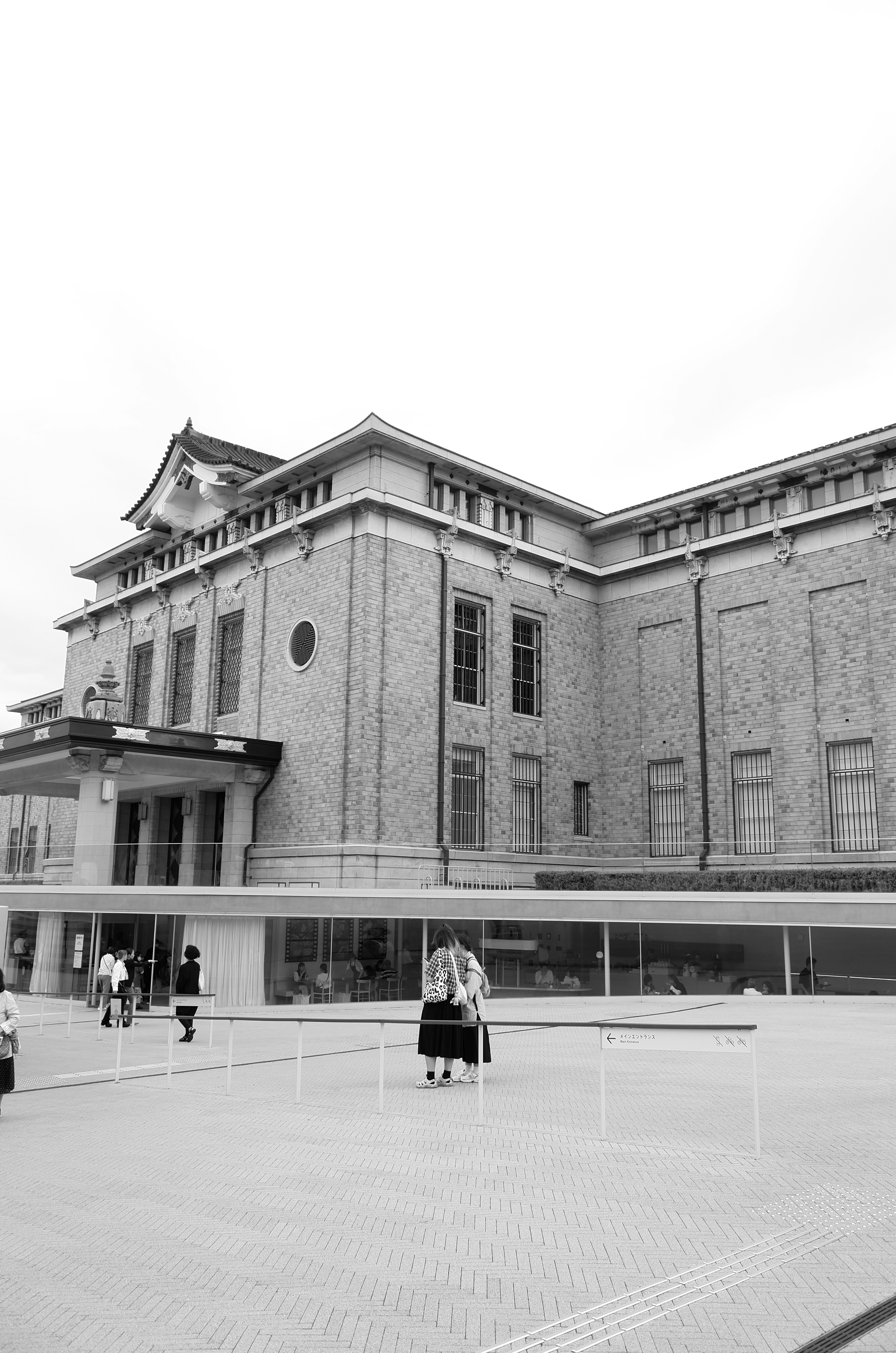 Black-and-white photograph of a brick building with tall windows and a glass entrance, set in a spacious plaza with a small group near the glass walkway.