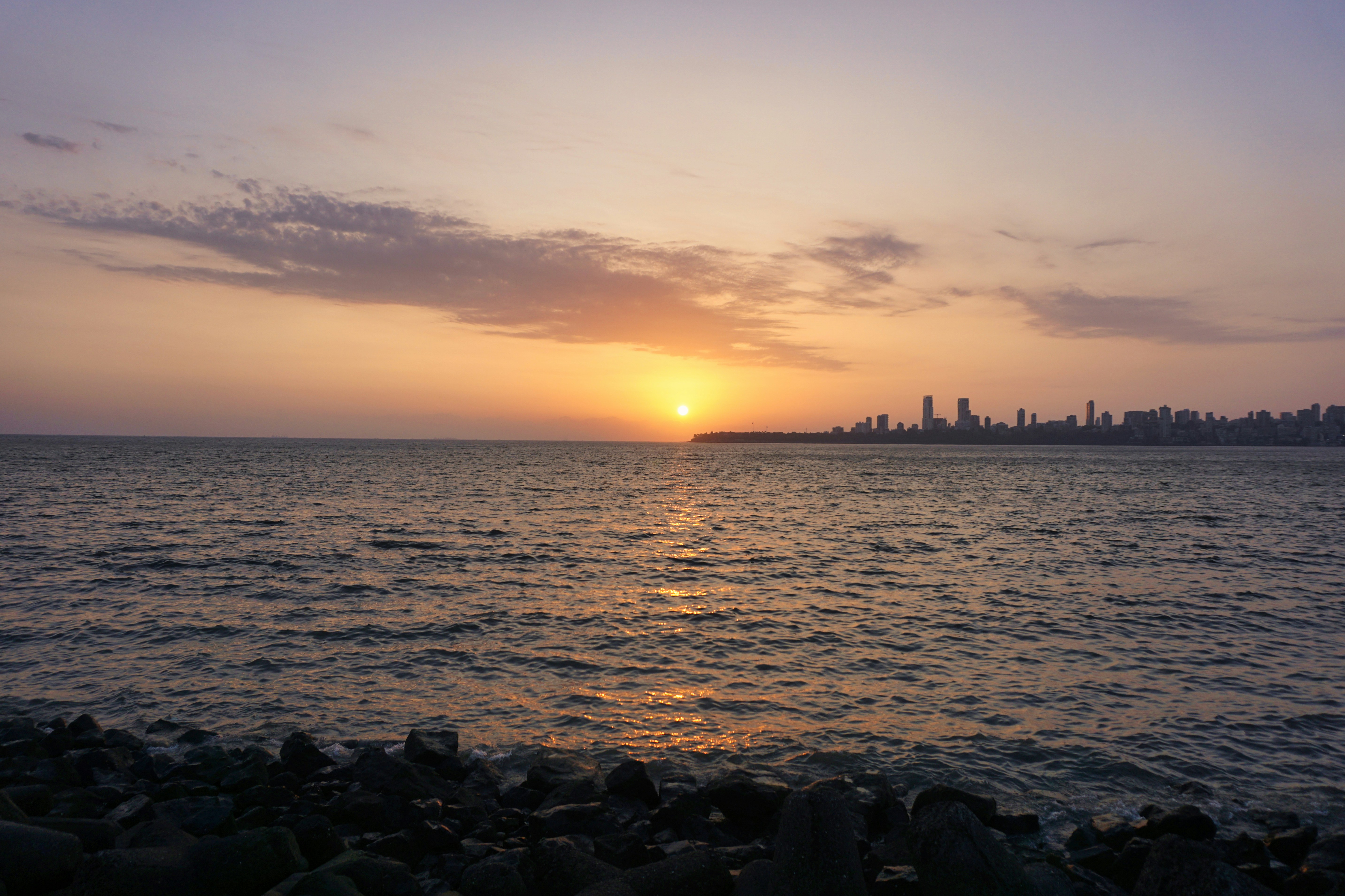 Sunset over a calm sea with a distant city skyline under a softly clouded sky.