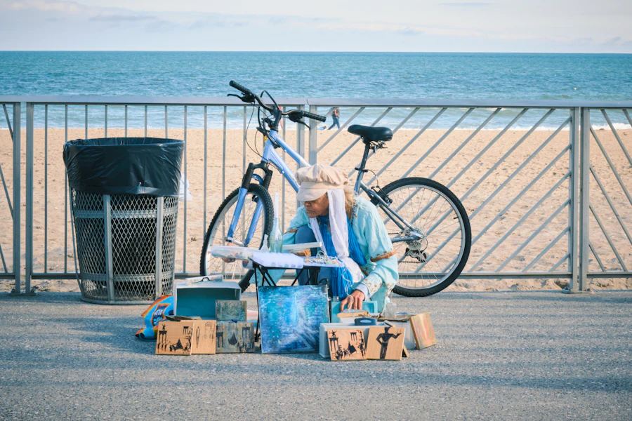A seated artist works among paints and small boxes on a seaside promenade, with a bicycle and railing framing the sandy beach and blue ocean behind. Soft daylight gives the scene a calm, coastal feel.