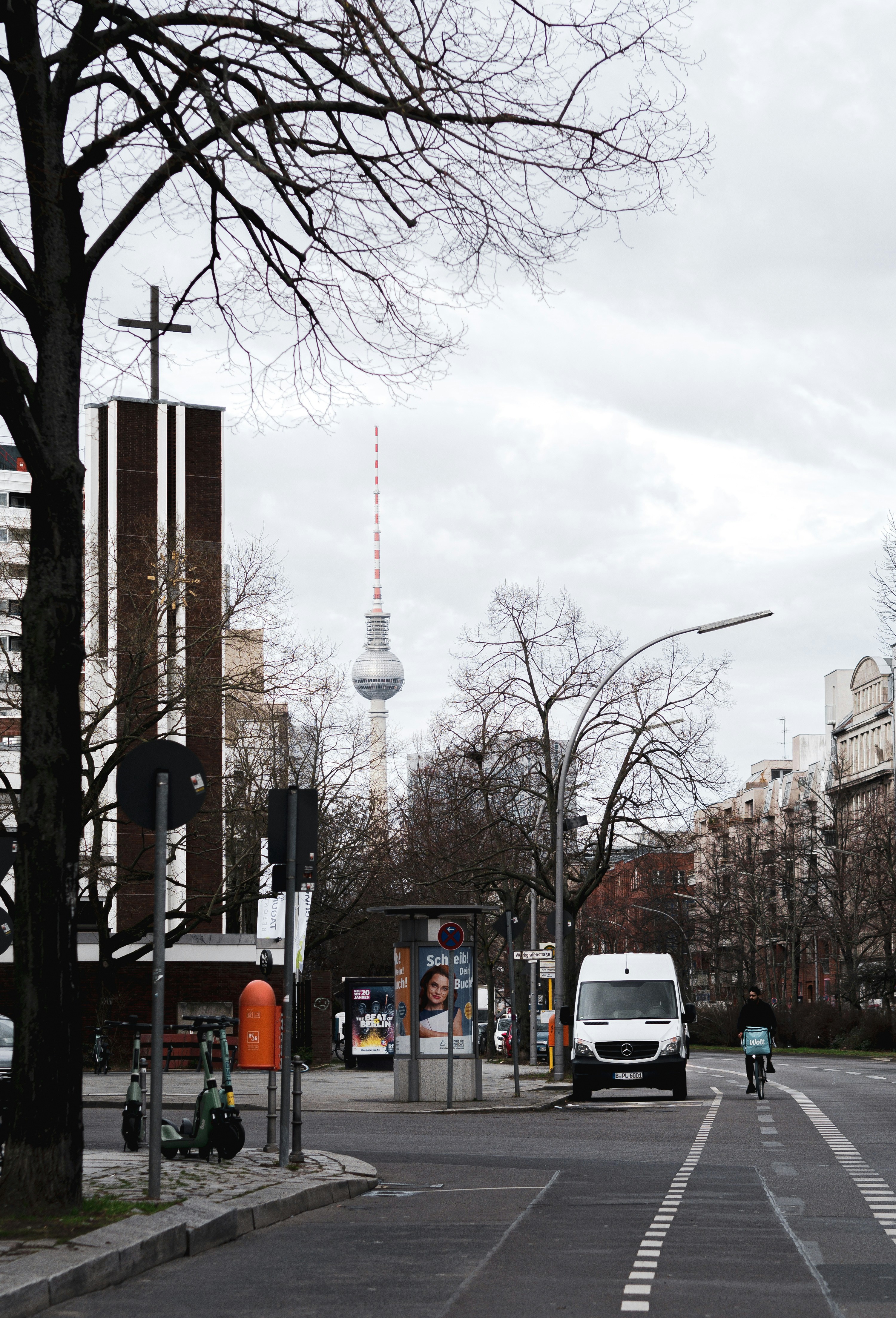 Berlin's iconic TV Tower looms over a quiet street scene, framed by bare trees and modern architecture. A cyclist navigates the urban landscape.
