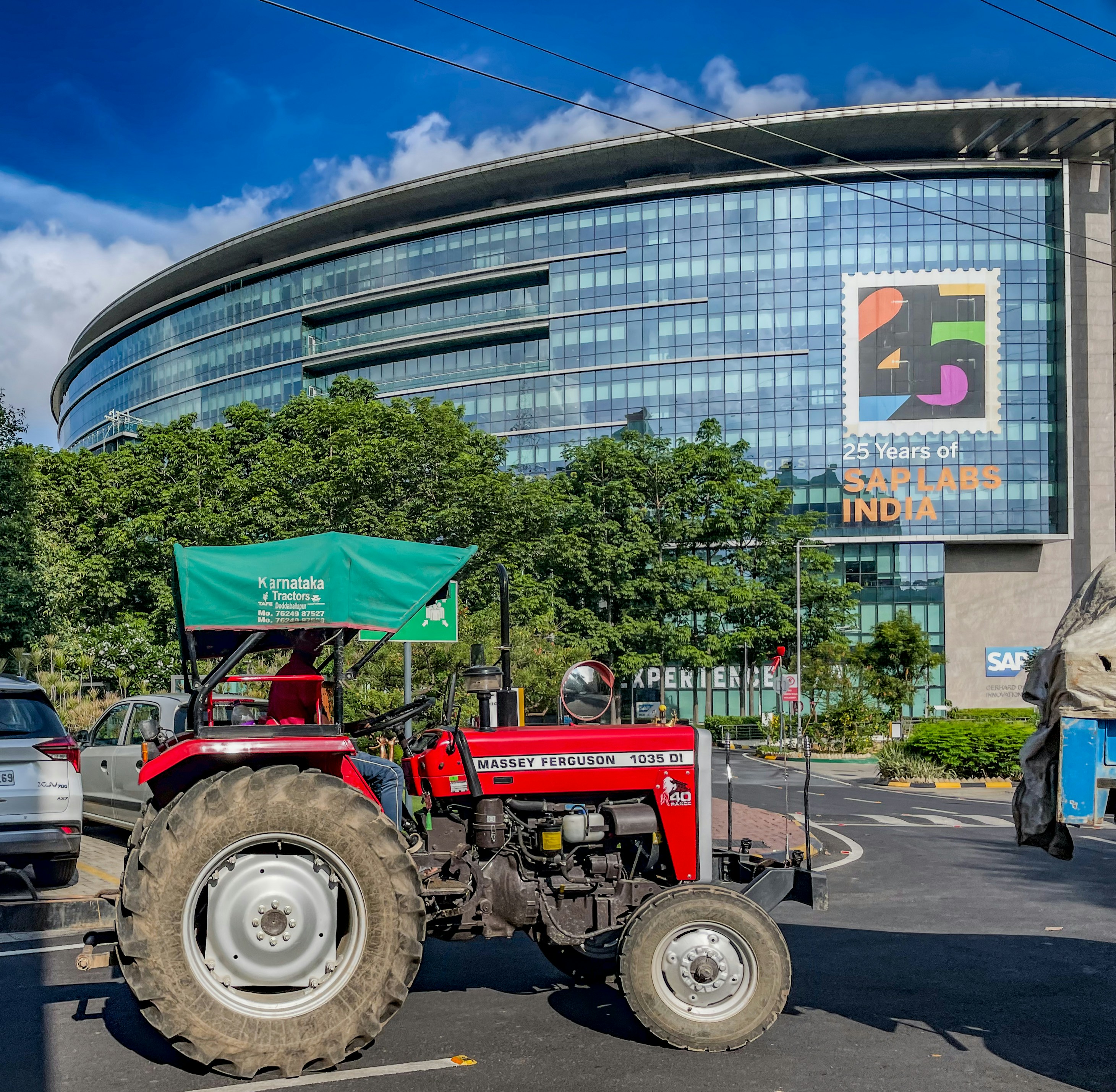 A red Massey Ferguson 1035 DI tractor is parked in front of the modern SAP Labs India building. The contemporary glass facade of the office contrasts with the rustic agricultural vehicle, highlighting a juxtaposition of rural and urban elements. The background features lush greenery and clear blue skies, adding to the vibrant and dynamic setting. The building's signage celebrates "25 Years of SAP Labs India," reflecting the intersection of technology and tradition in this urban landscape.