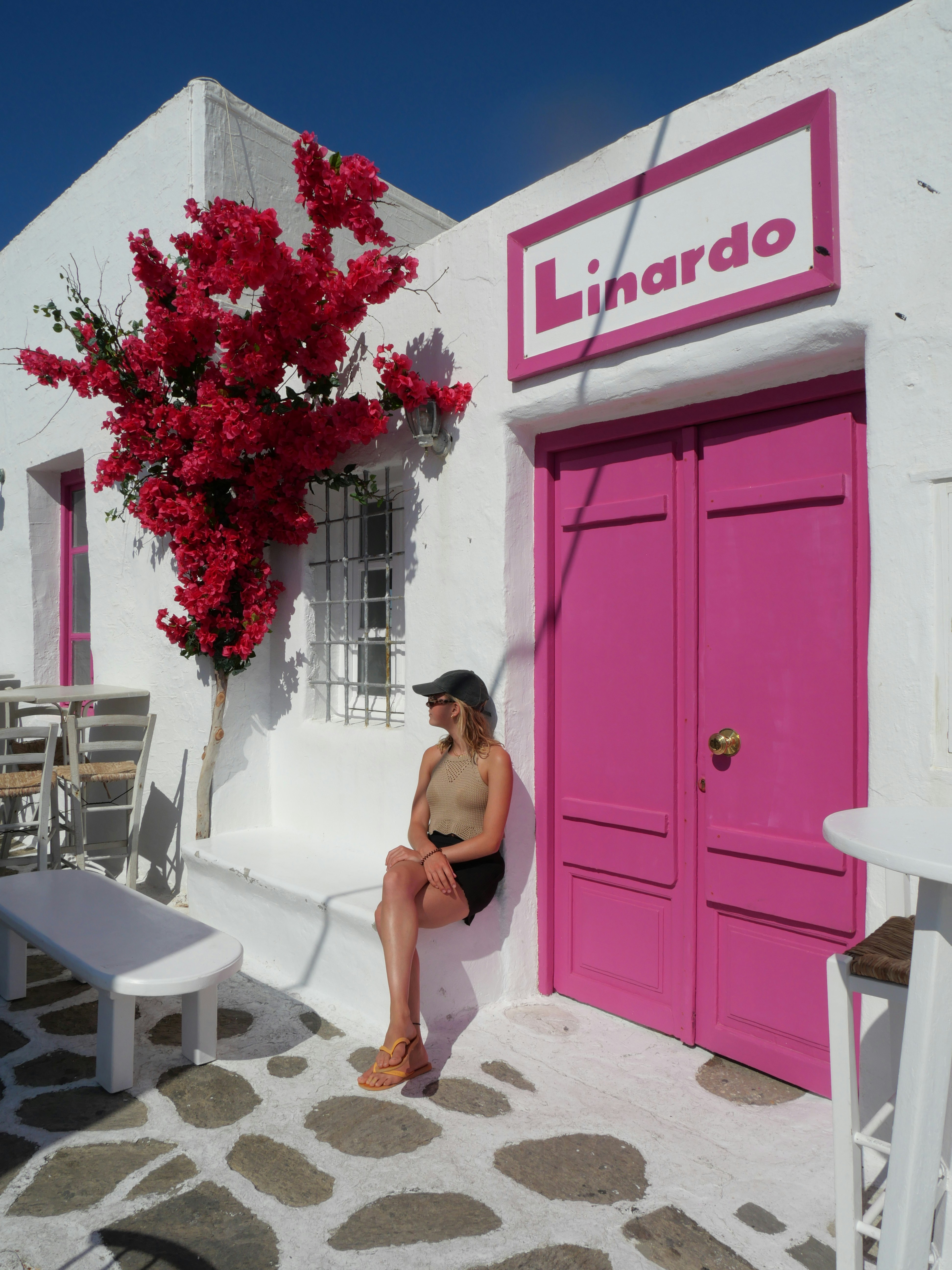 A woman sits casually near a vivid pink door as bougainvillea blooms against a white wall. The scene conveys a sunlit, Mediterranean courtyard mood.