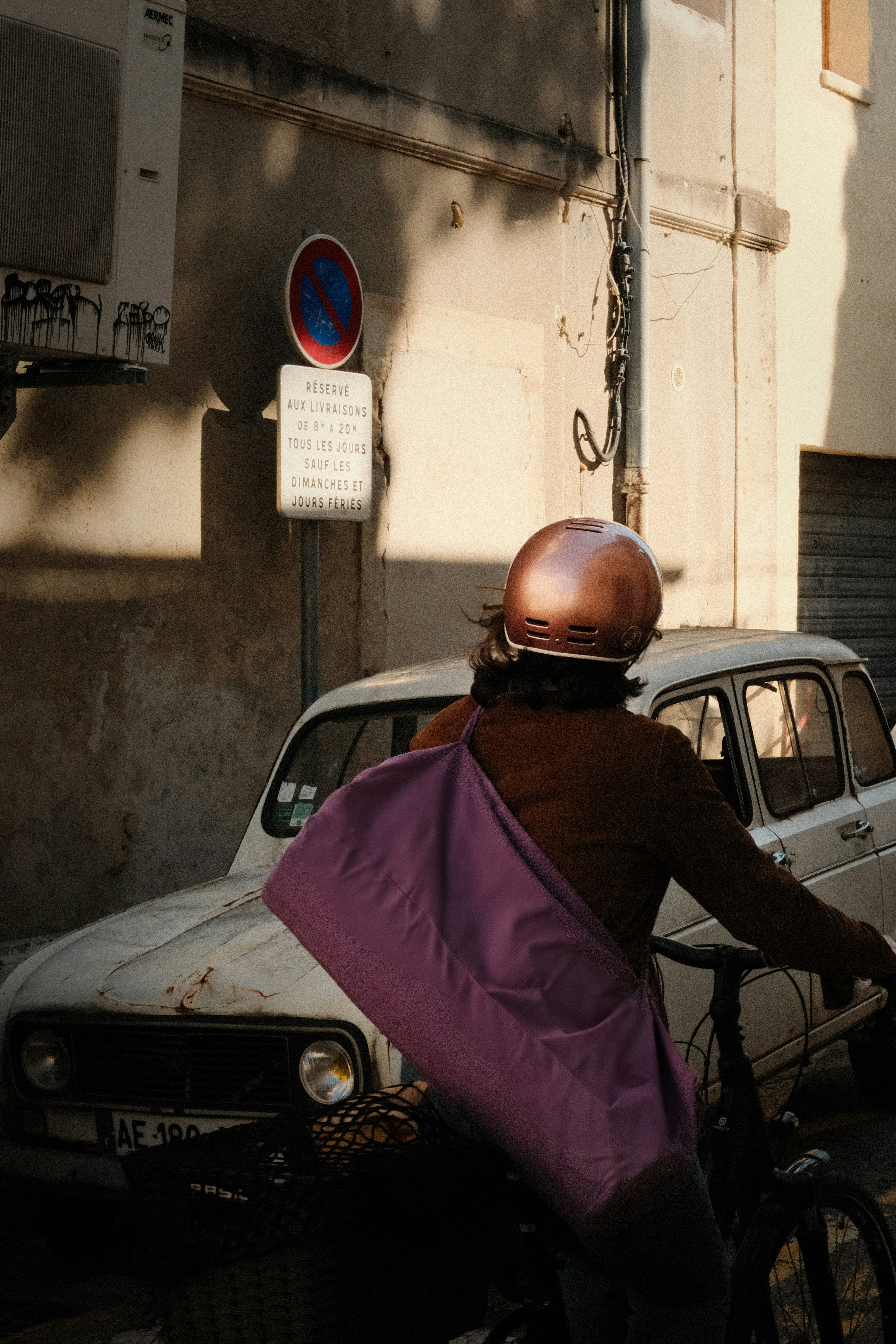 Cyclist with a purple bag rides past a vintage car on a sunlit street.