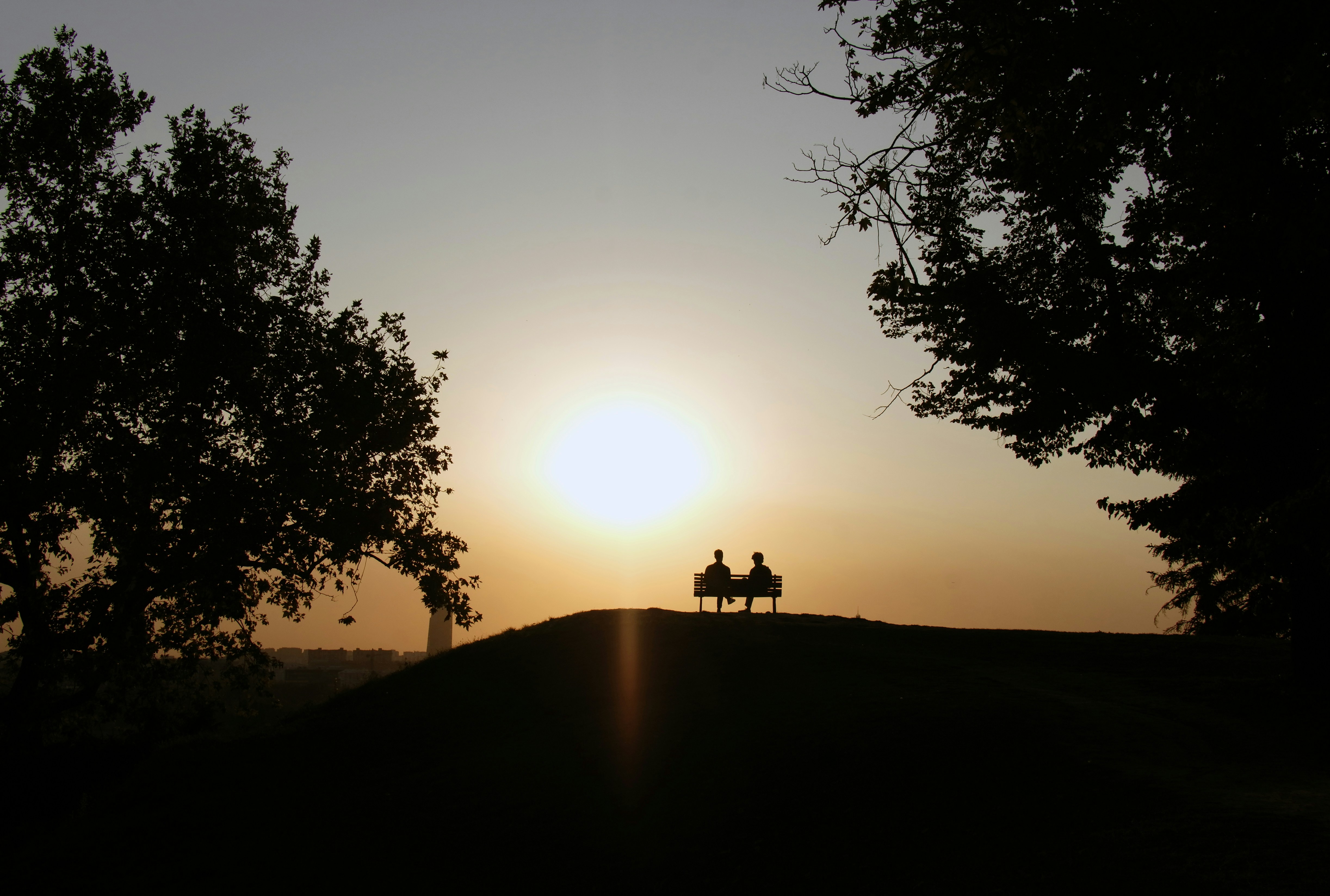 Two figures seated on a hilltop, silhouetted against a glowing sunset, framed by trees. The scene evokes a sense of tranquility and connection.