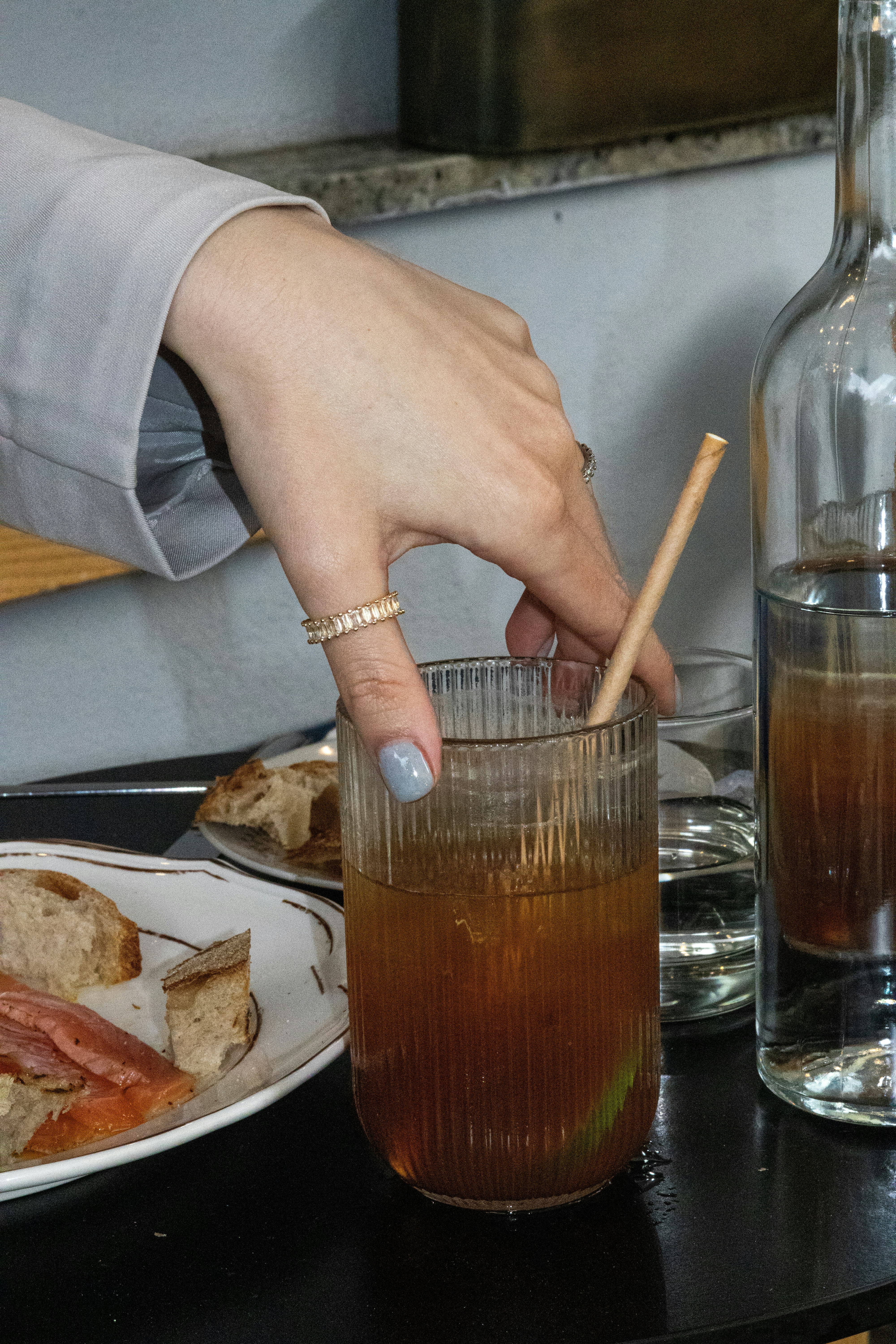 A hand stirs an amber-colored drink in a ribbed glass with a straw at a dining table, with bread and shrimp on a plate and a bottle in the background.