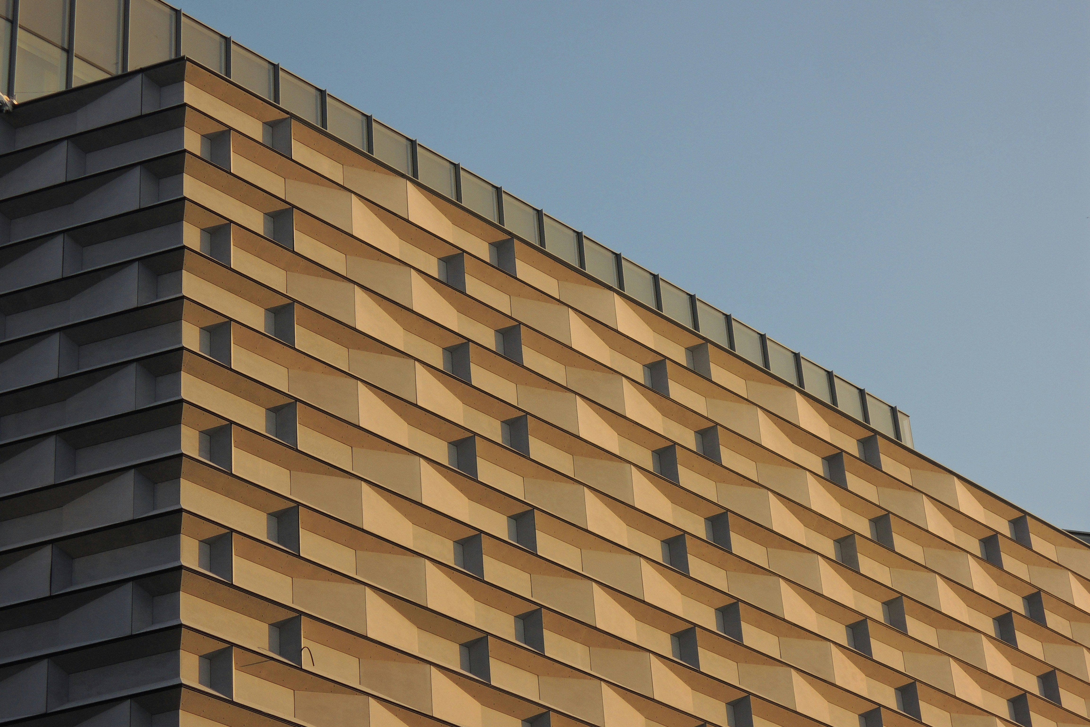 Golden-toned facade of a modern building featuring diagonal sunlit panels and a repeating grid of small windows.