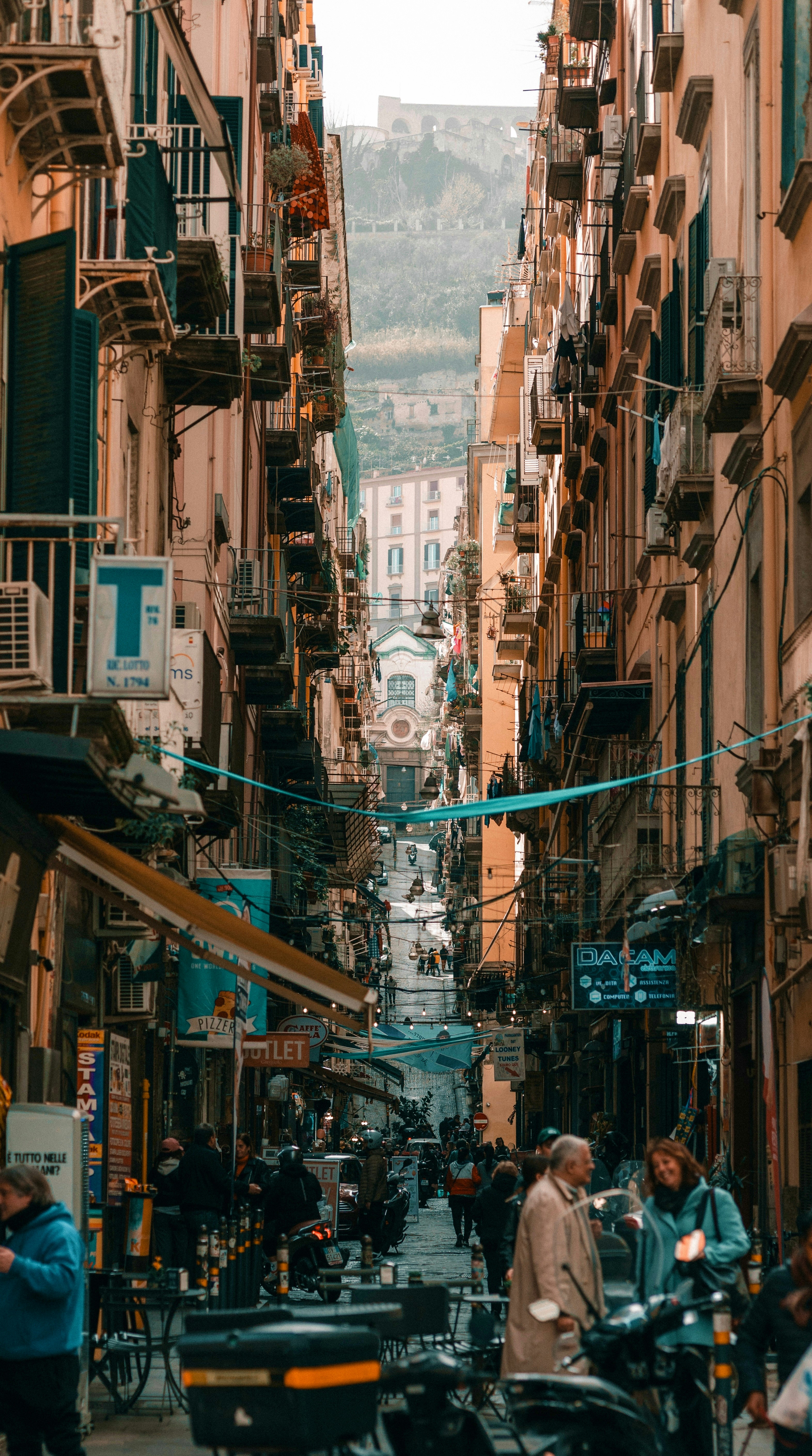 Narrow street bustling with people and lined with balconies, revealing a glimpse of a distant hillside and architecture.