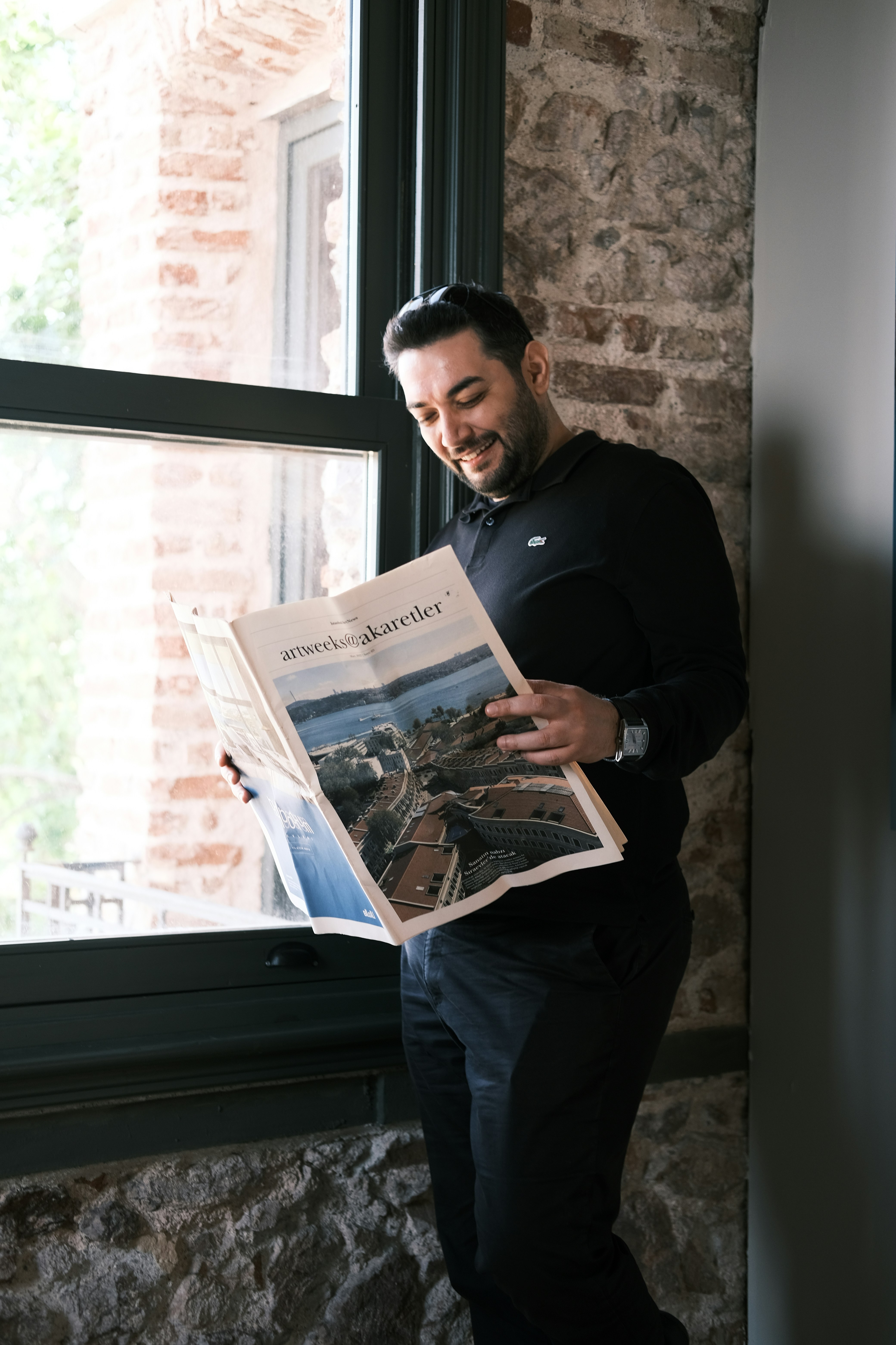 Man standing by a window, reading a large book with a smile, natural light illuminating a rustic brick wall.
