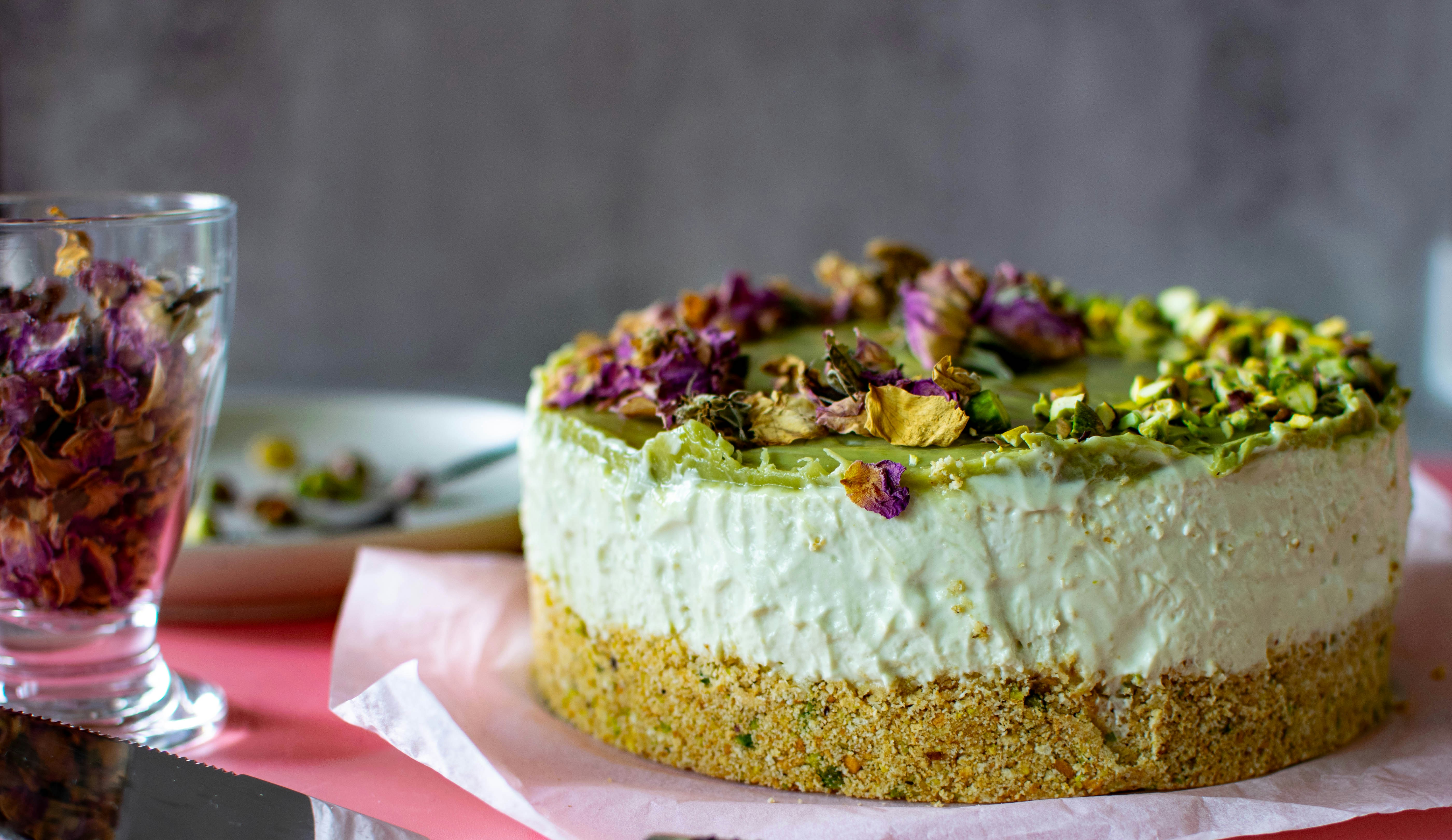 A beautifully decorated cake topped with pistachios and dried rose petals, set against a soft, blurred background. The scene is complemented by a glass of dried flowers nearby.