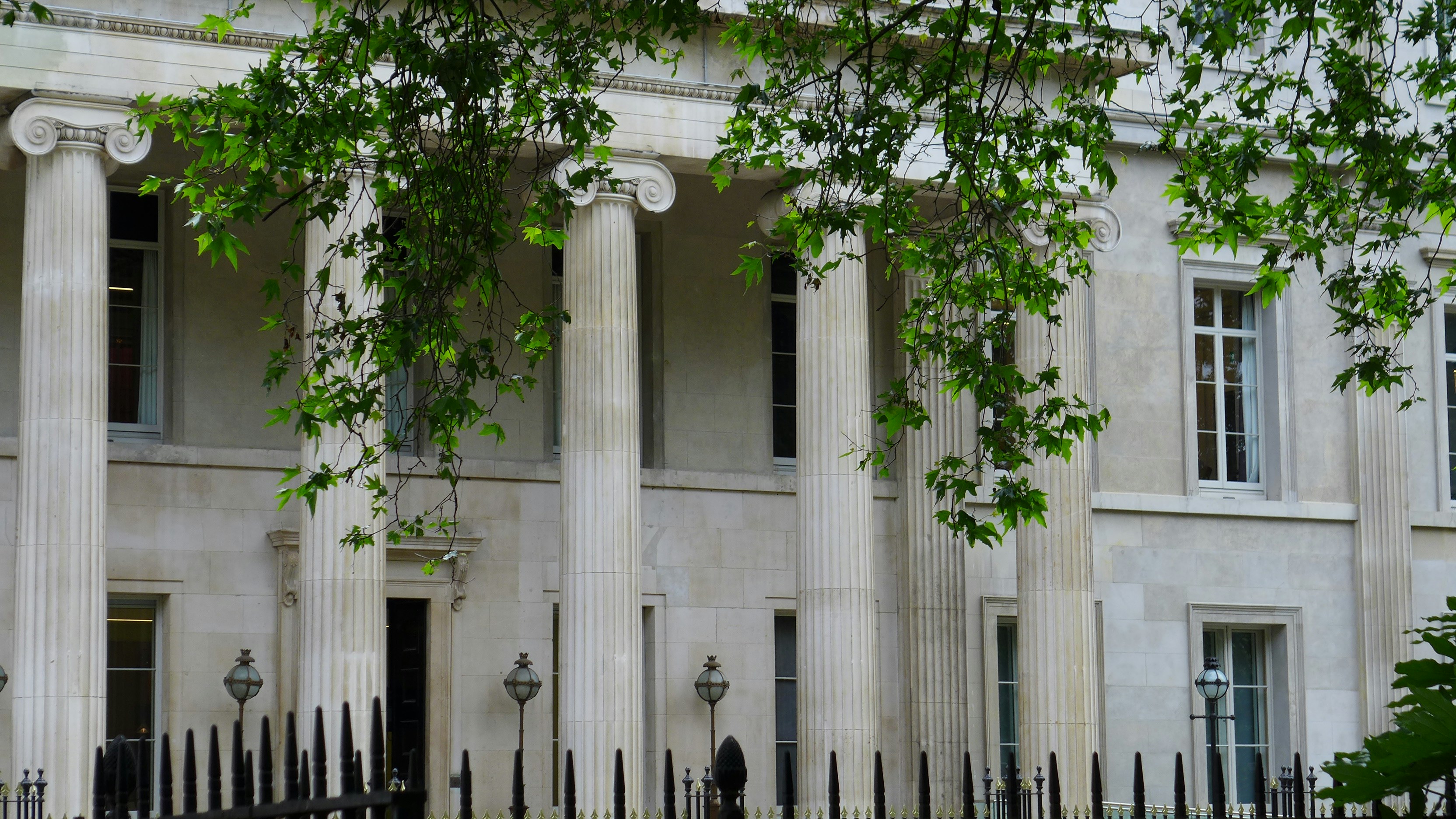 Neoclassical building facade with tall columns and a wrought-iron fence, framed by overhanging green leaves.