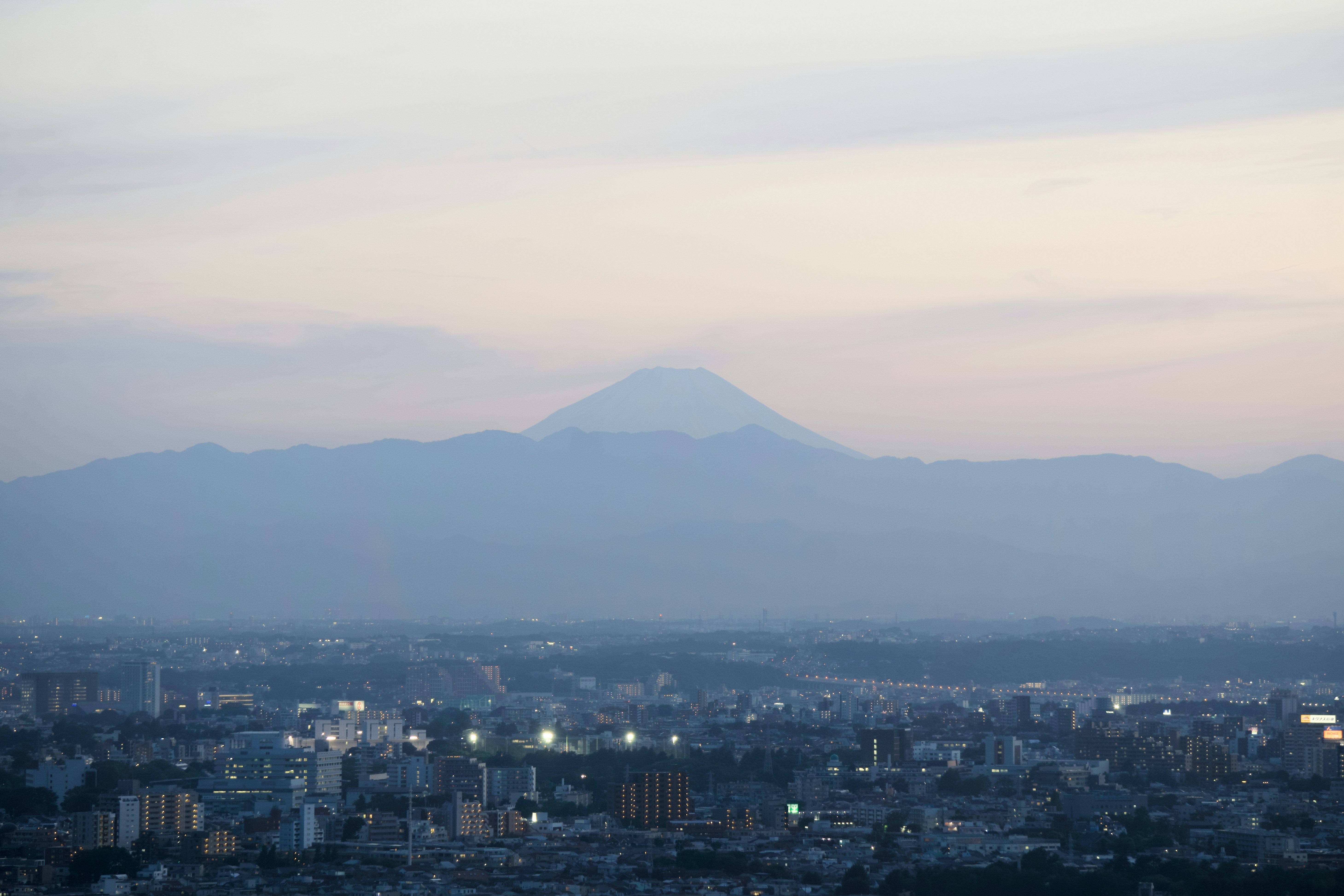 Softly illuminated silhouette of Mount Fuji rising above a sprawling cityscape during twilight.