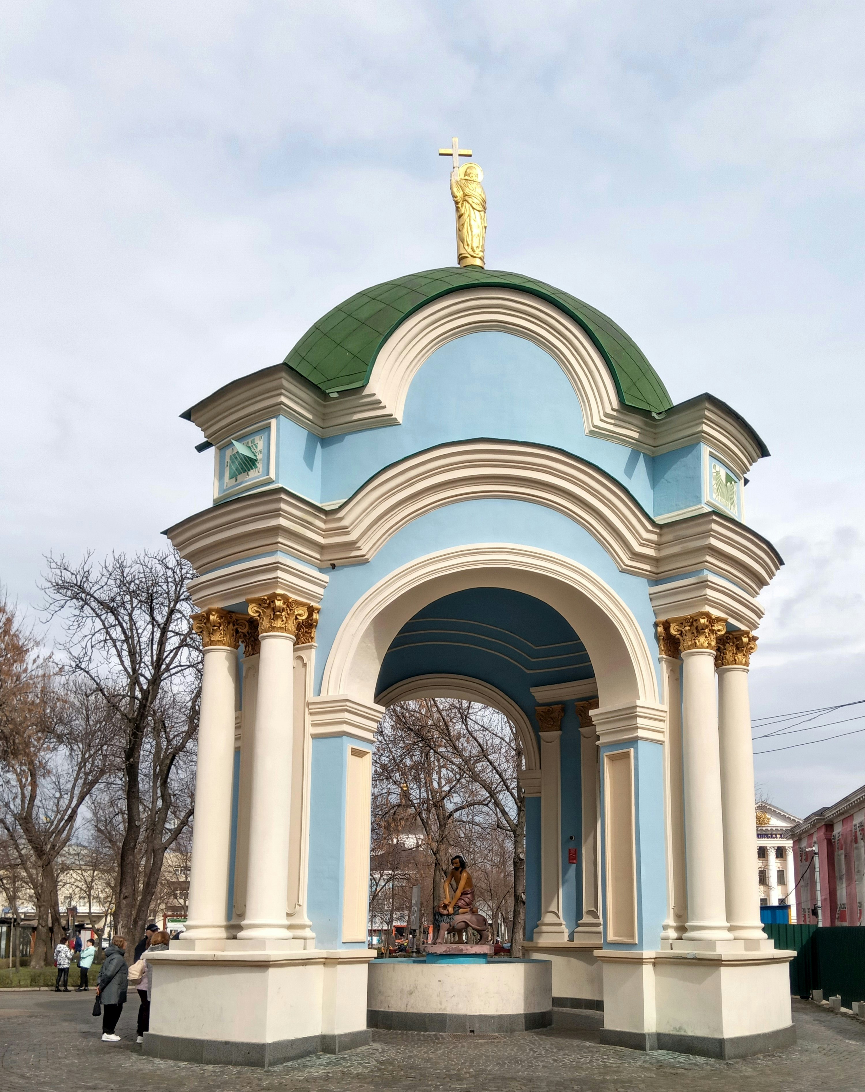 Photograph of a cerulean neoclassical arch with white columns and a green domed roof, crowned by a golden statue, in a sunlit plaza.