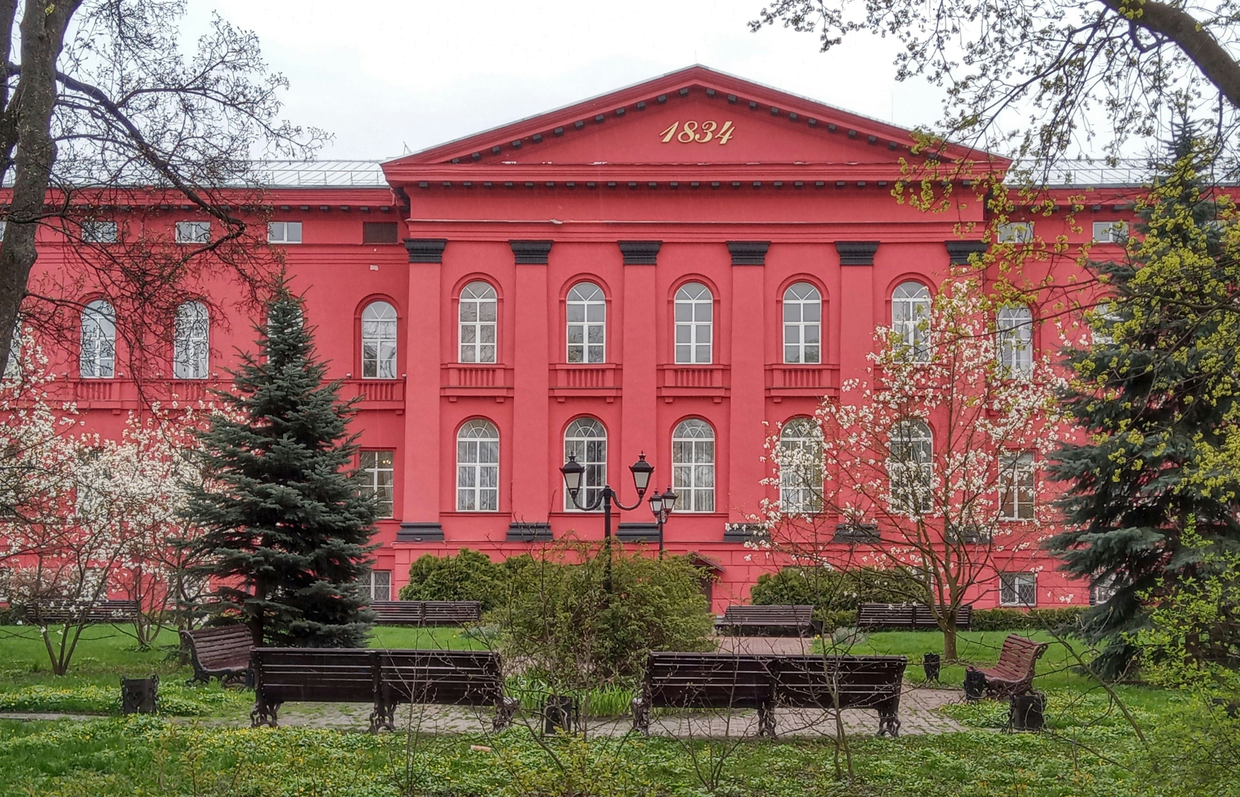 Red-brick neoclassical facade with a prominent pediment dated 1834, centered in a park setting. Park benches, trees, and a lamp post populate the foreground.