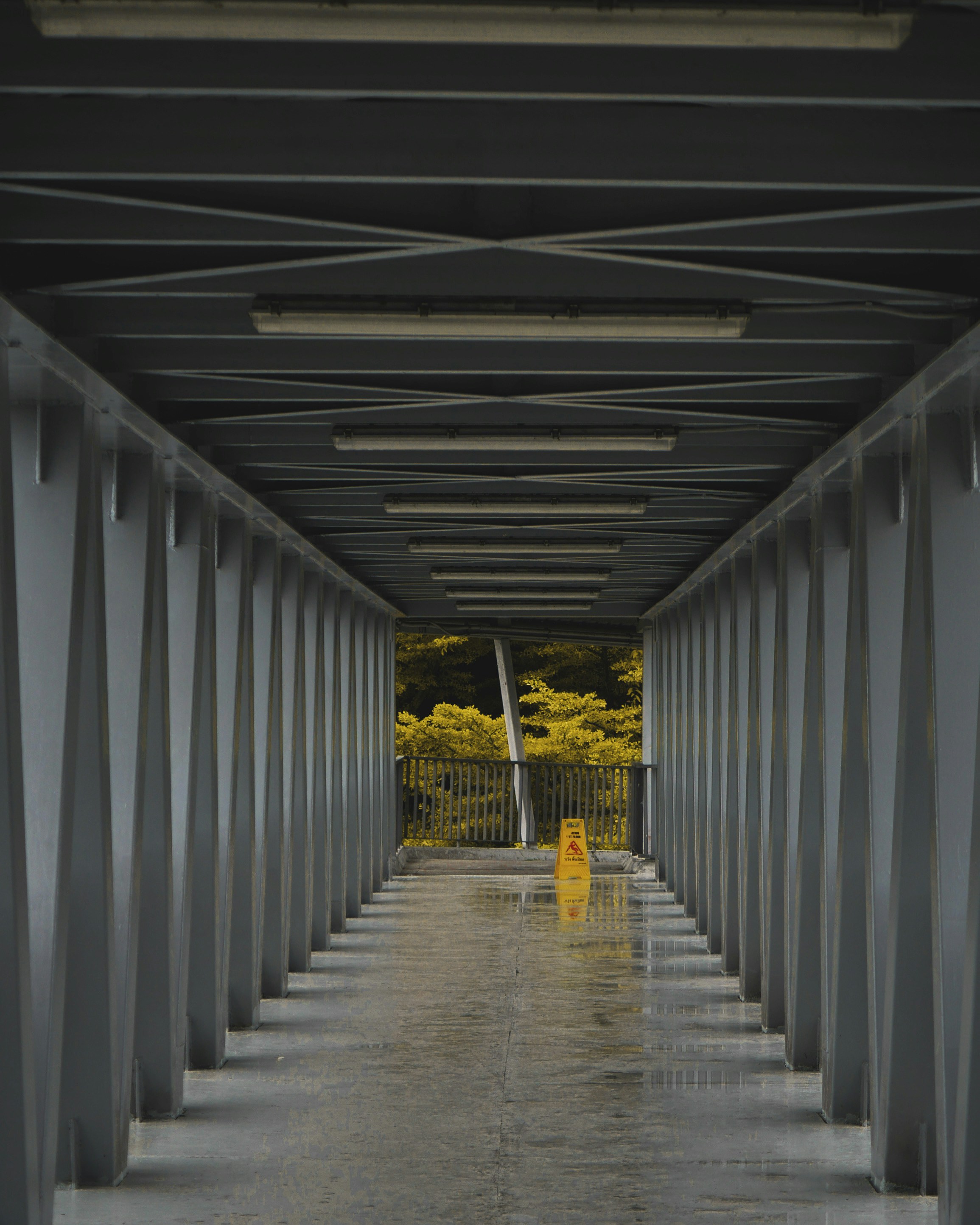 Long, wet covered steel corridor creates a tunnel effect with repeating vertical supports receding toward a sunlit green clearing. A caution sign sits on the slick floor near the exit, adding a splash of color and context.
