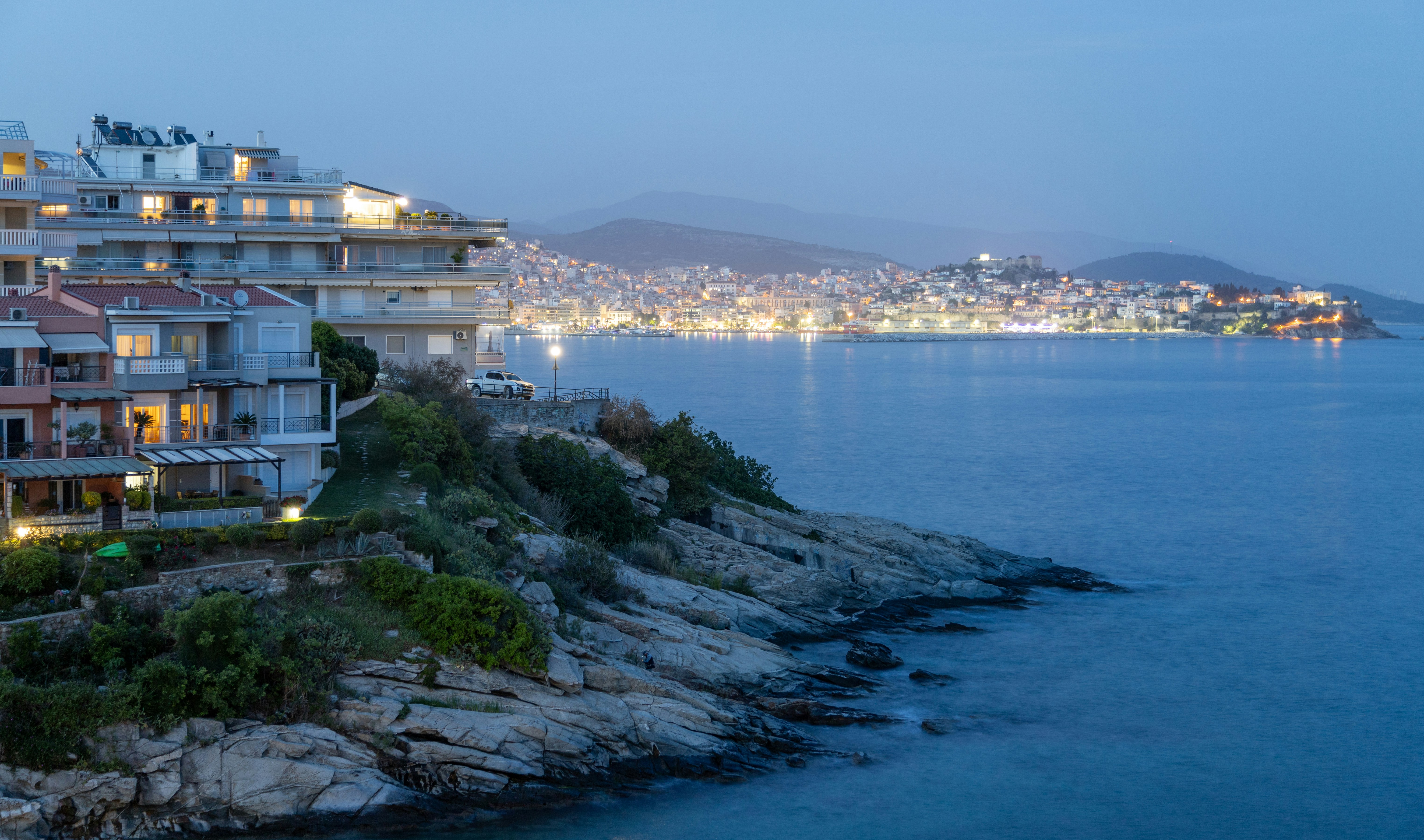 Coastal buildings overlooking a calm sea at dusk with distant city lights.
