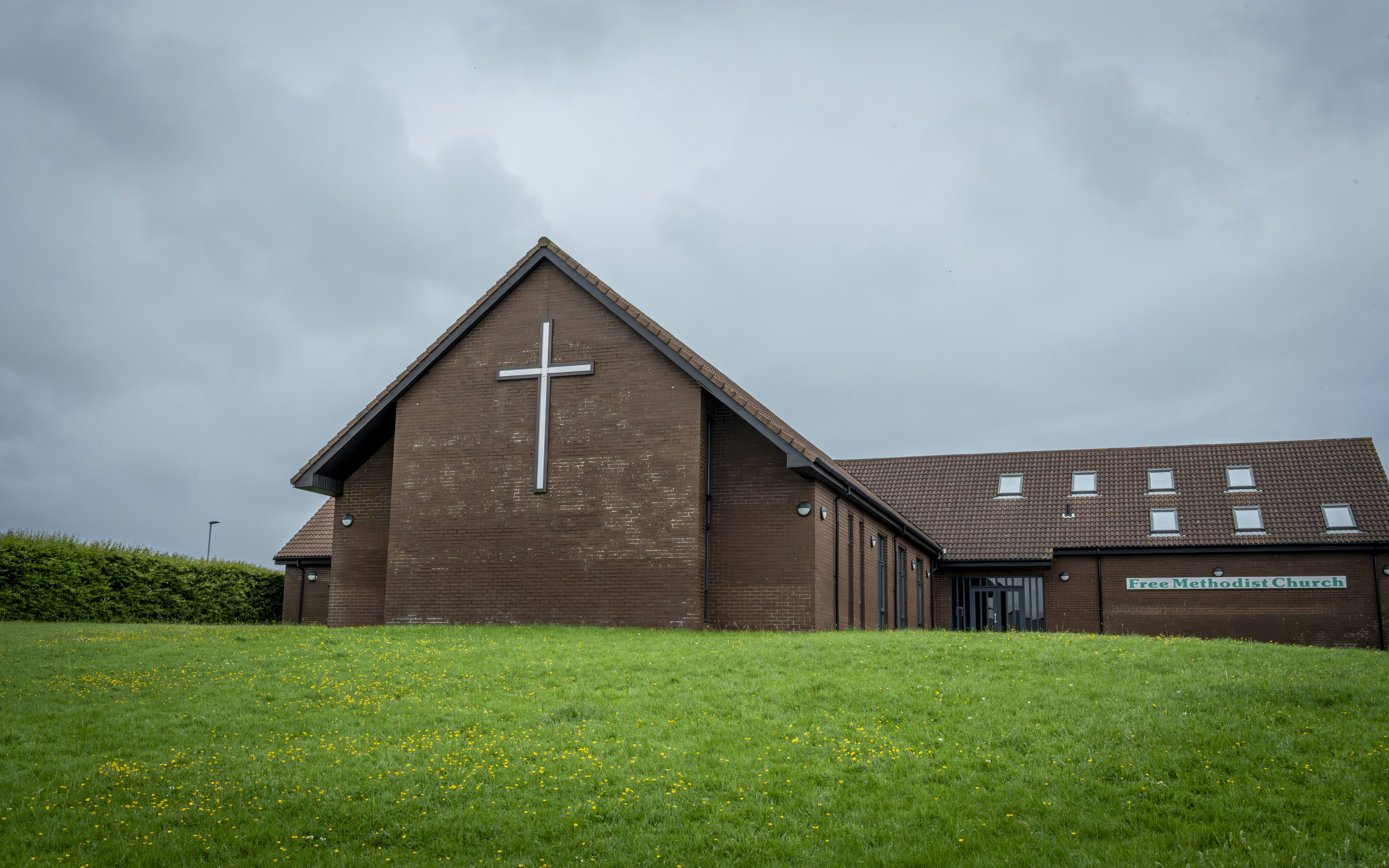 A contemporary church building with a prominent cross on its façade, set against a backdrop of overcast skies and lush green grass.