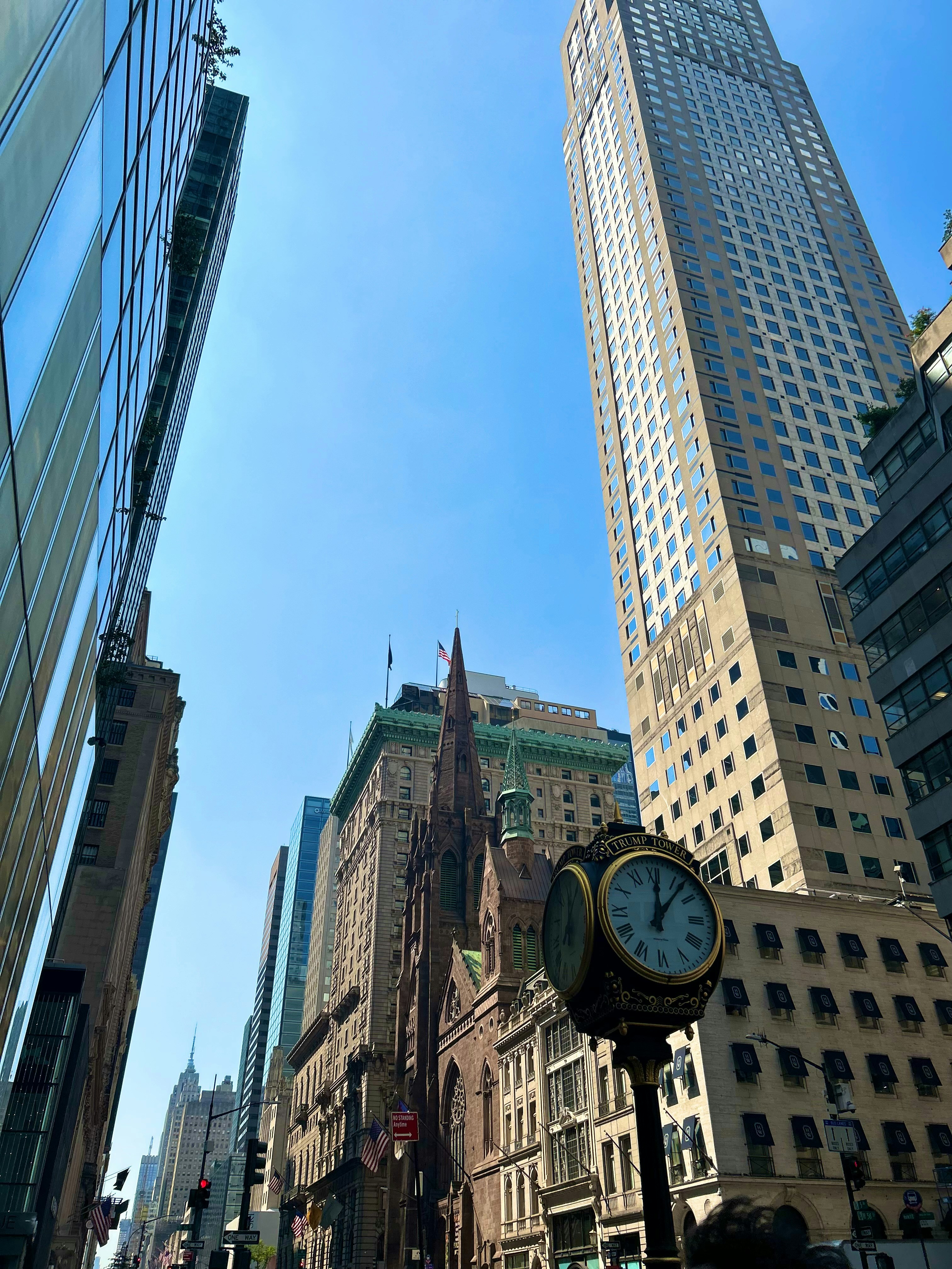 Historic clock stands in front of a blend of architectural styles, showcasing the juxtaposition of old and modern buildings against a clear blue sky.