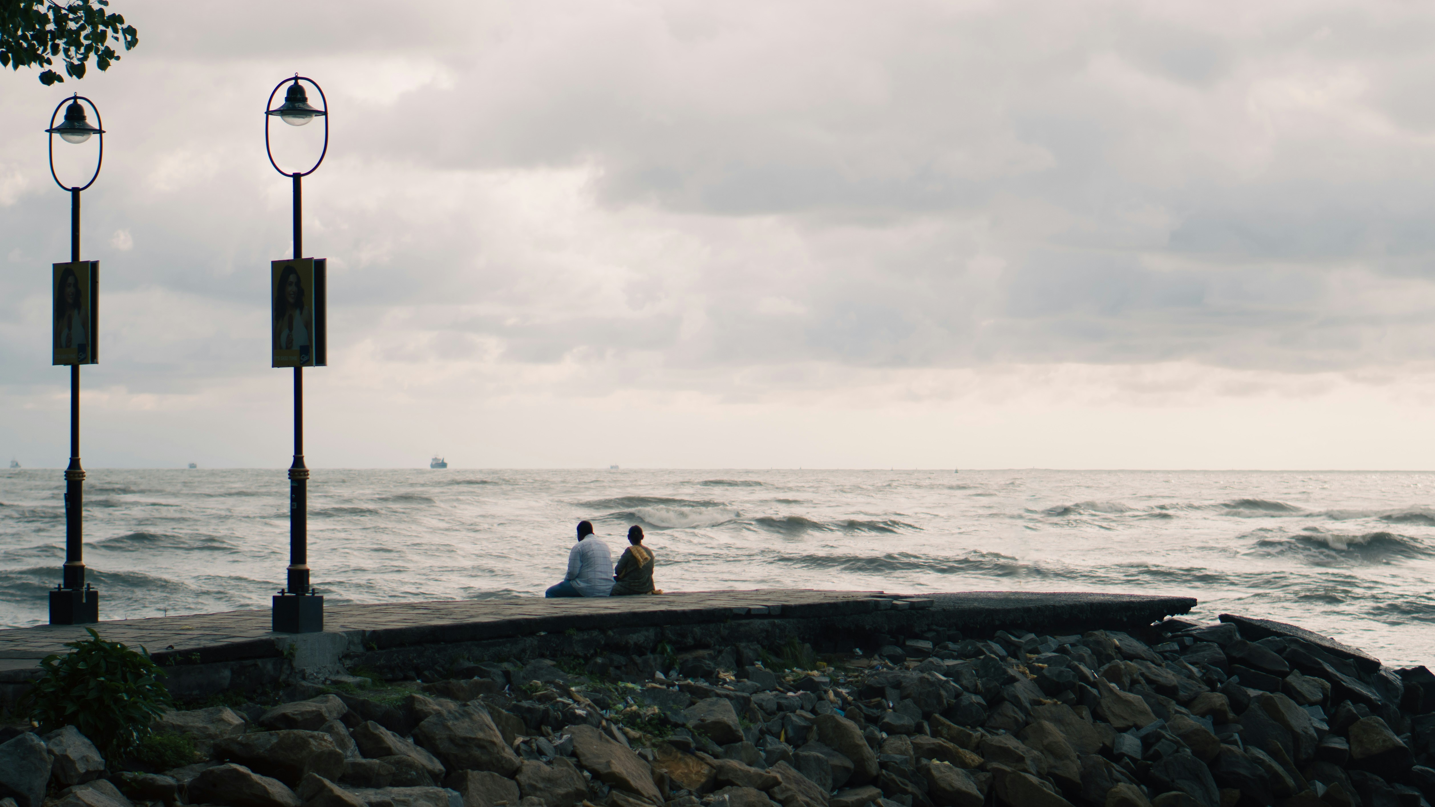 Two figures seated on a rocky shore, gazing at the expansive sea under a cloudy sky. Streetlamps frame the scene, adding a touch of urban charm.