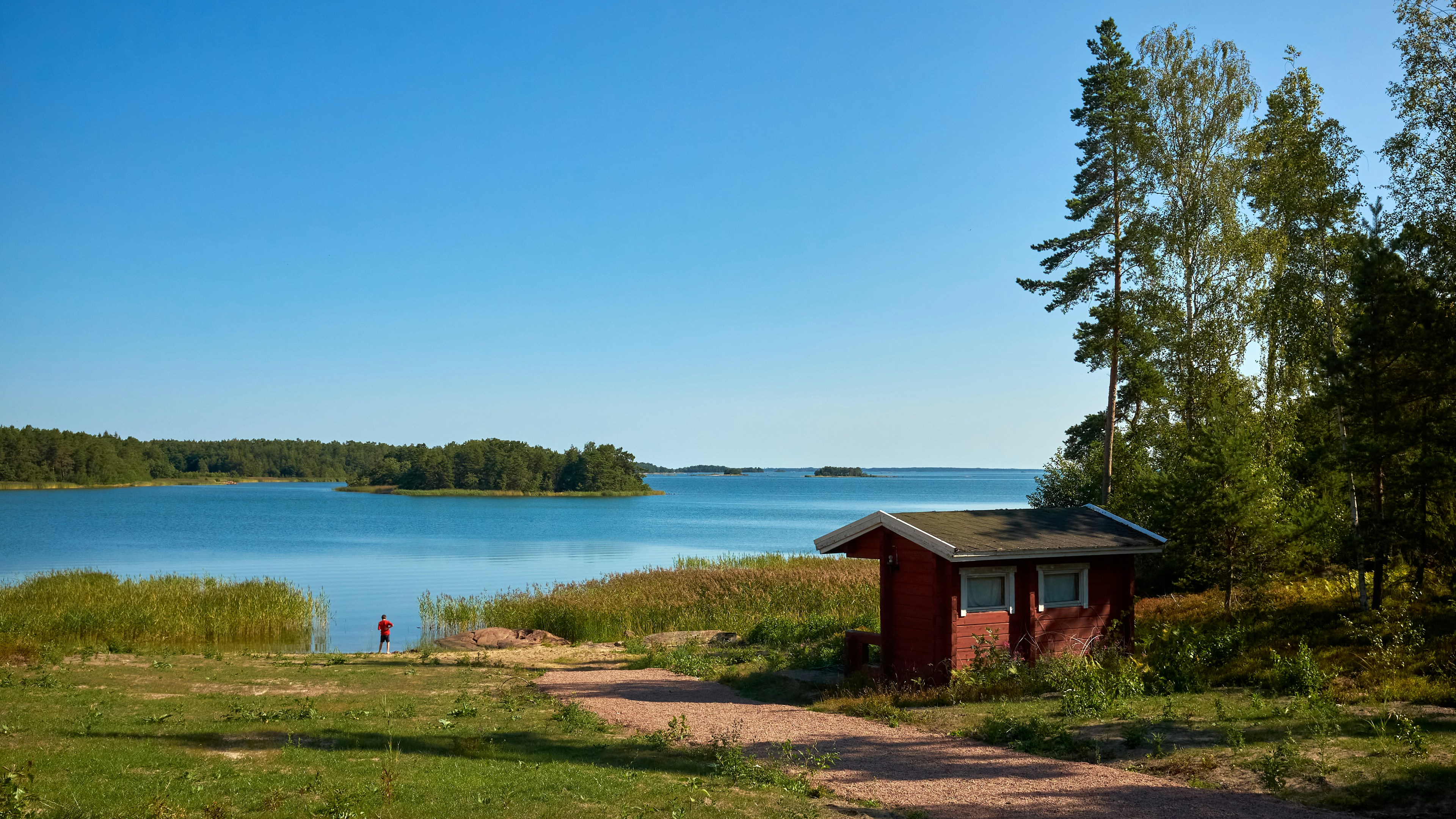 Rustic cabin by a serene lake under a clear blue sky, surrounded by lush greenery.