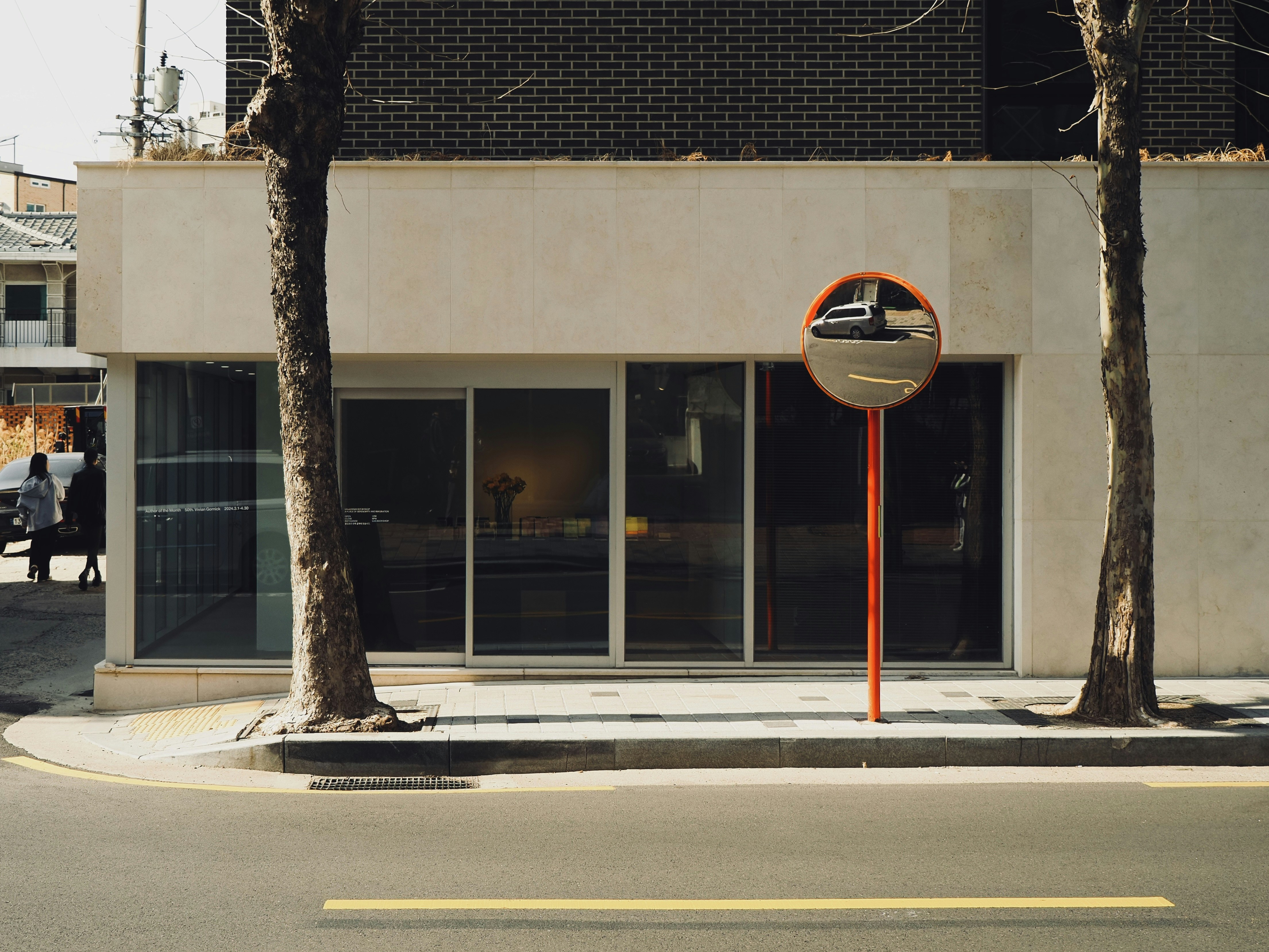Round convex mirror on a red post reflects the street-facing storefront beneath two trees. Minimalist glass panels and calm urban rhythm define the scene.