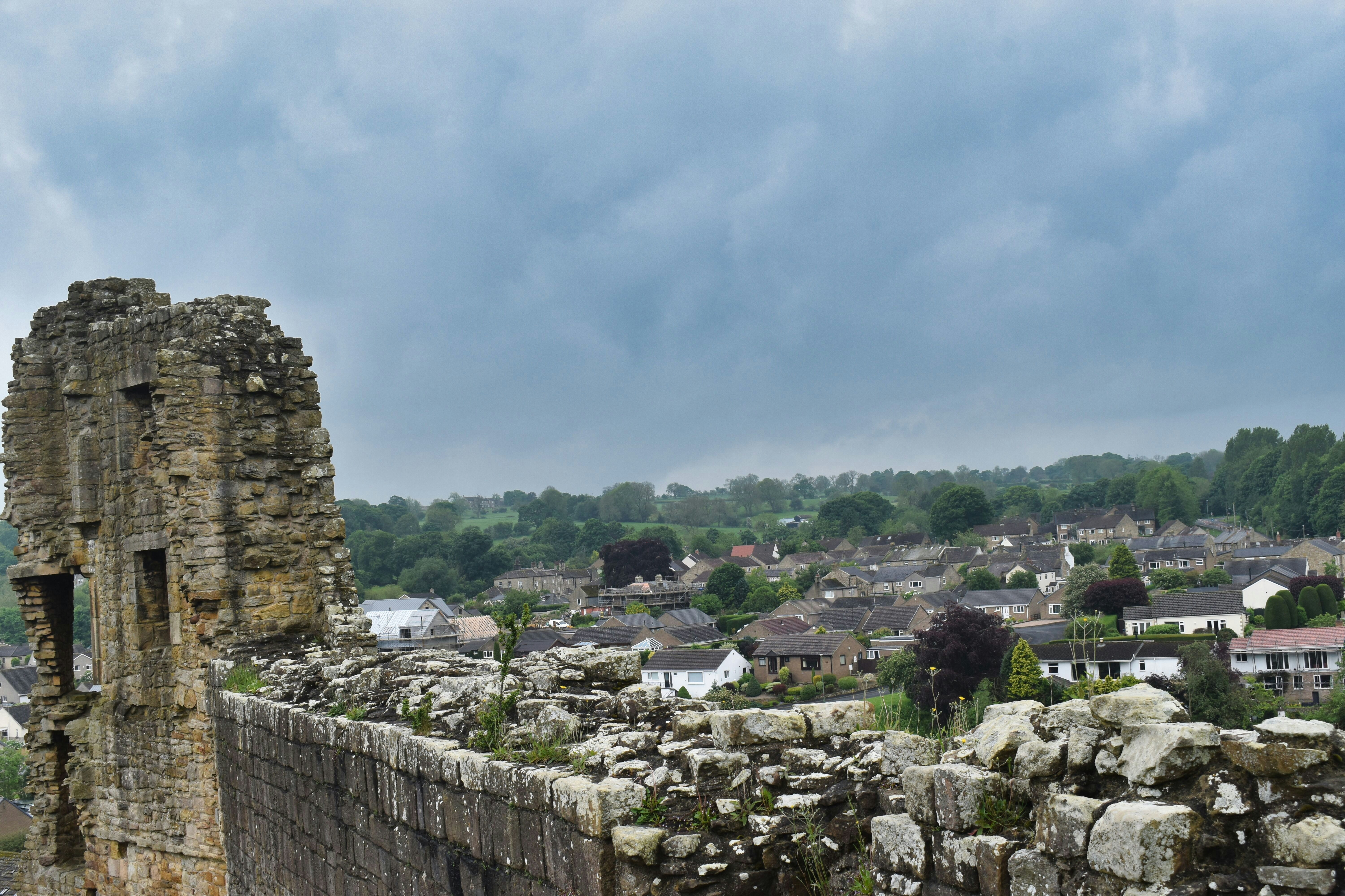 Ancient stone ruins rise above a quaint village, contrasting with the lush green landscape and overcast sky. The scene captures the blend of history and contemporary life.