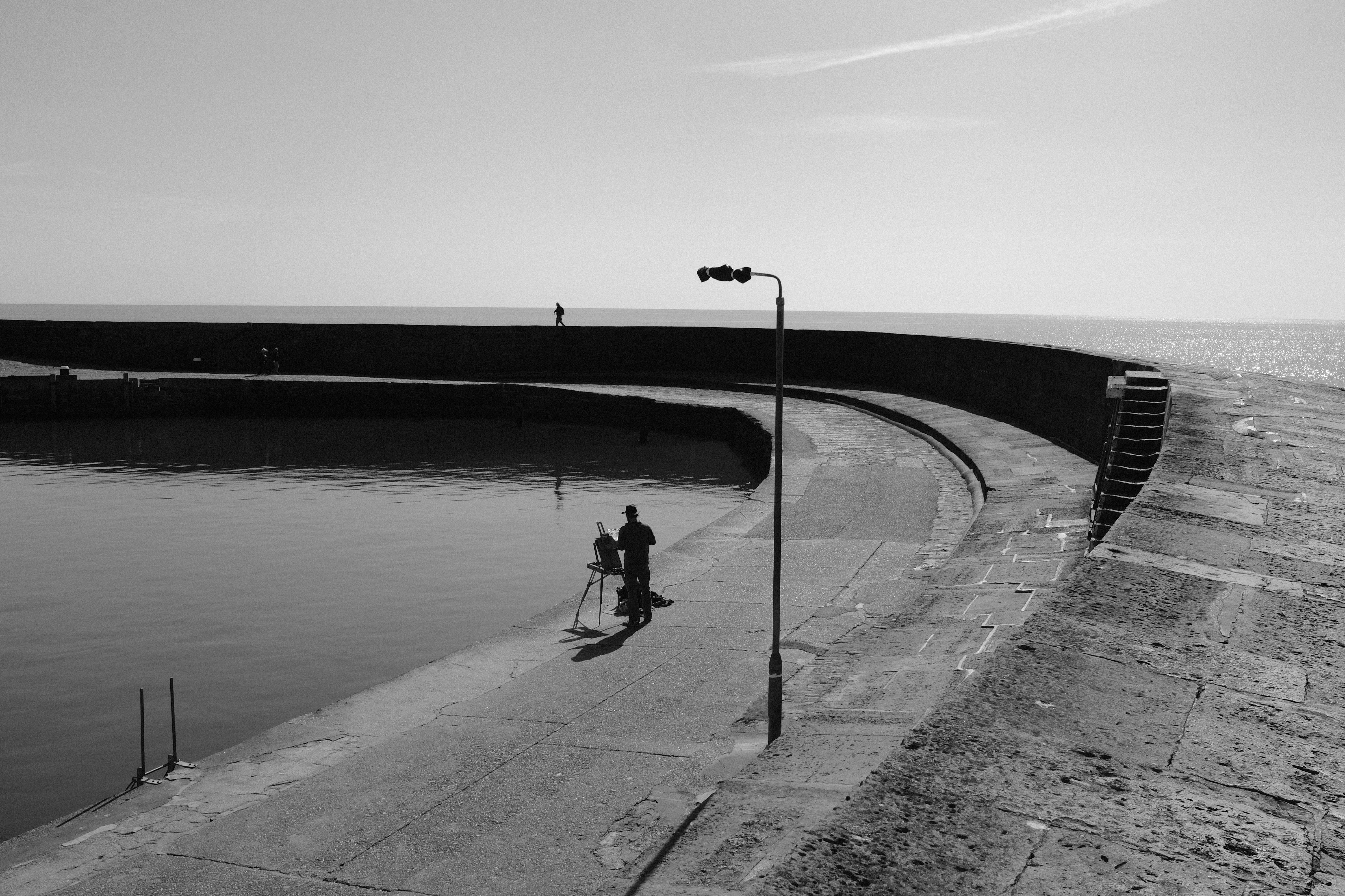 Black and white view of a solitary figure walking along a curved seawall under a clear sky.