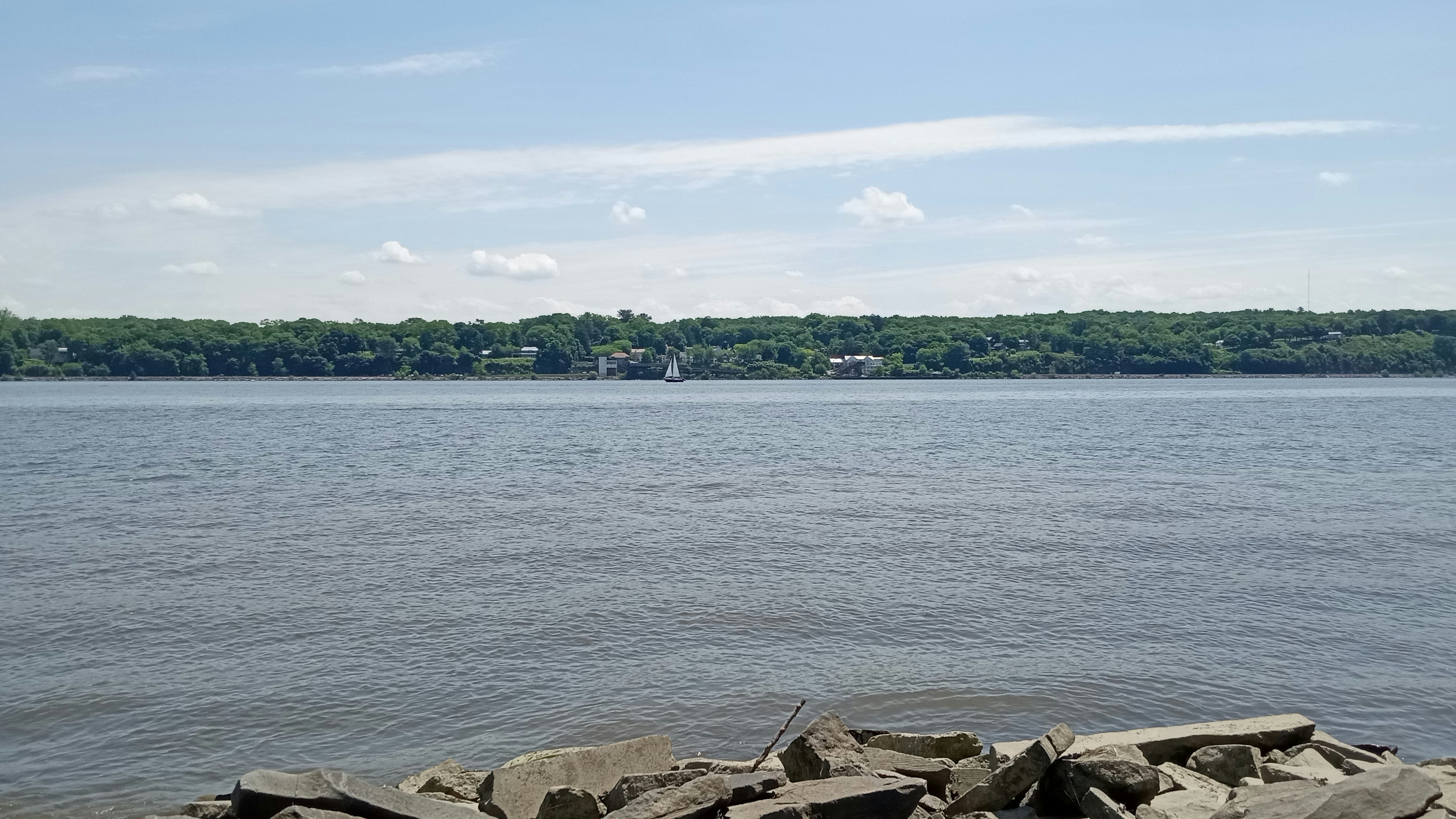 Calm lake scene with a rocky foreground and tree-lined far shore. A small sailboat marks the horizon under a clear blue sky.