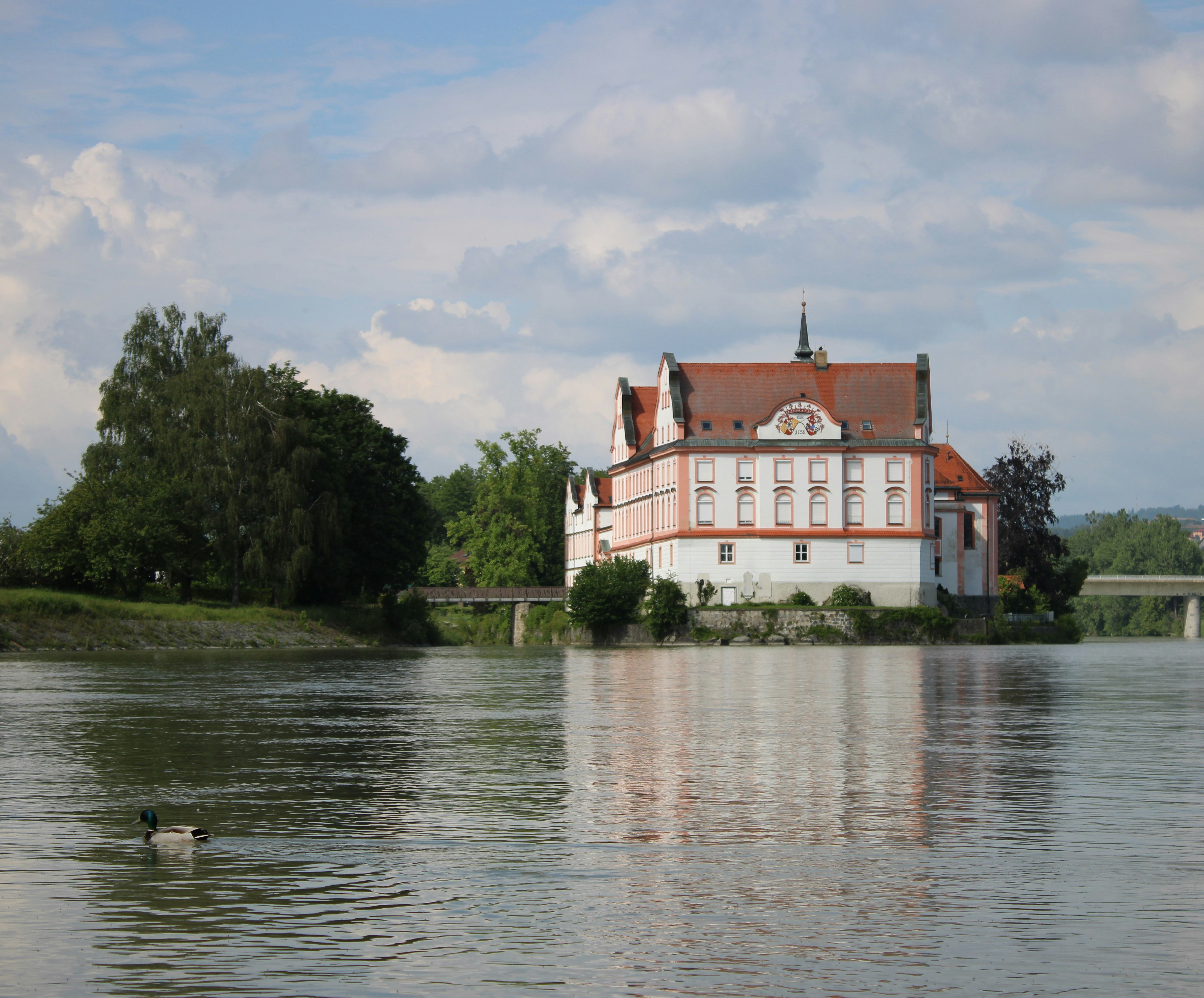 Historic chateau with red roof reflecting on calm river waters under a partly cloudy sky.