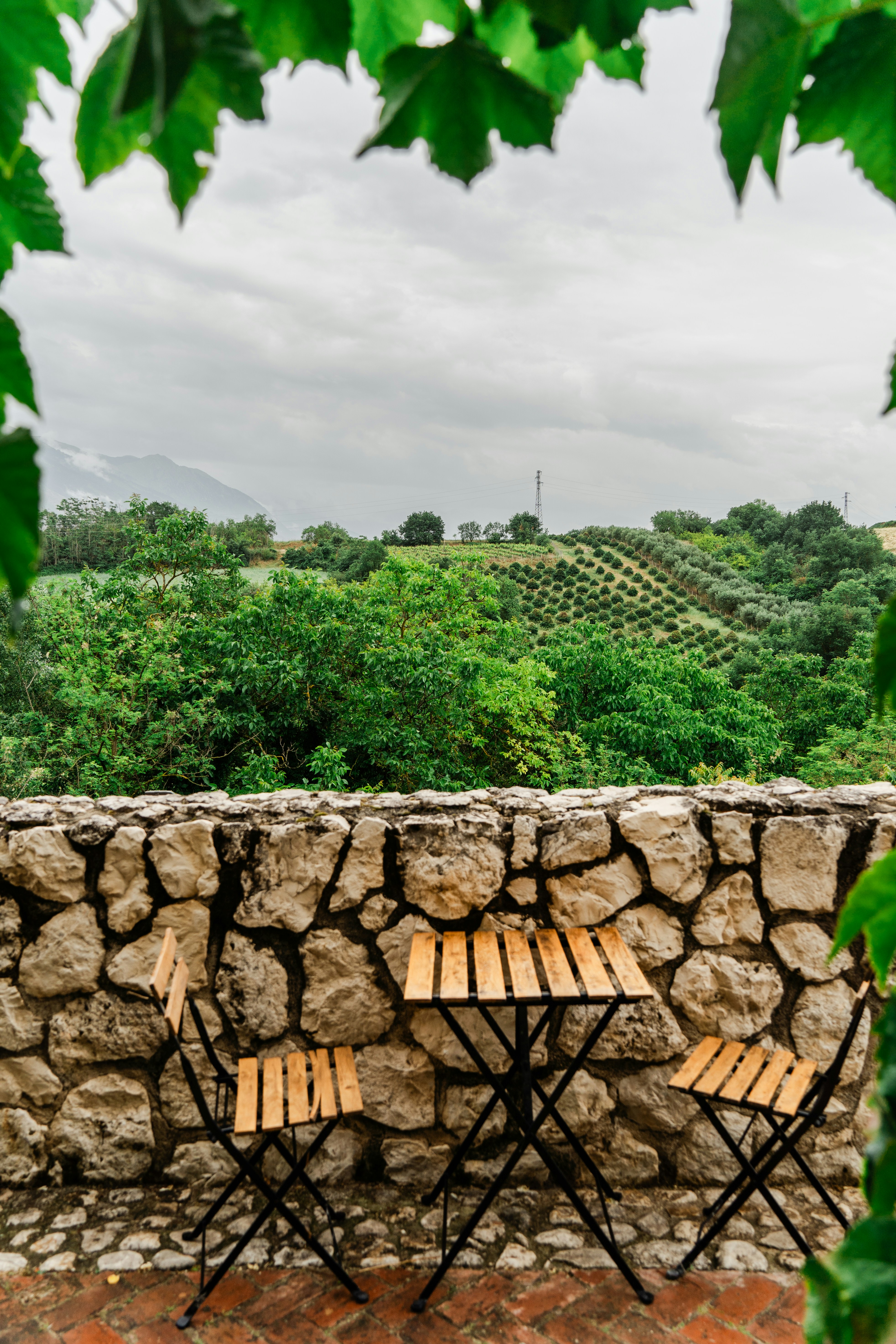 Wooden bistro table and chairs set against a rustic stone wall, framed by vibrant green leaves, with rolling hills and trees in the background.