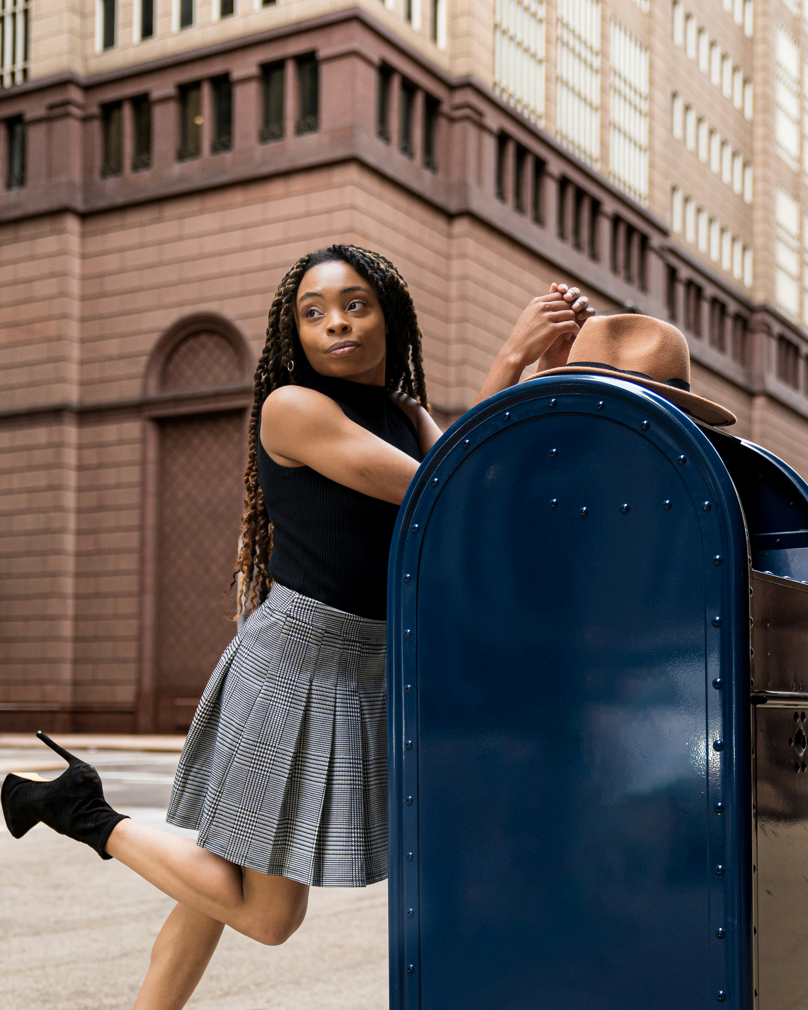 A woman in a sleeveless black top and plaid skirt poses mid-step beside a large blue mailbox against a brick urban backdrop.