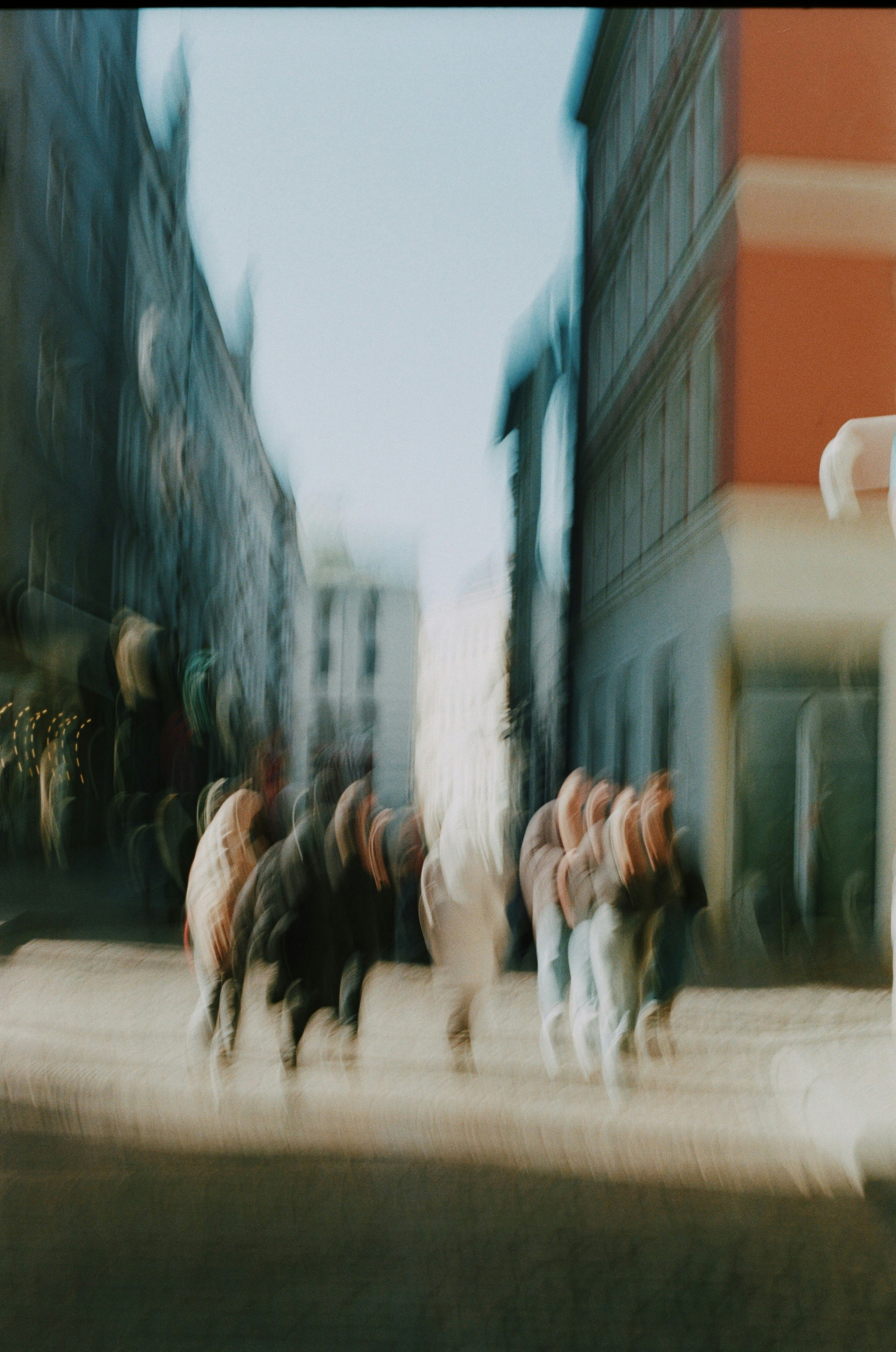 A motion-blurred city street with indistinct pedestrians forming soft, elongated silhouettes between buildings.