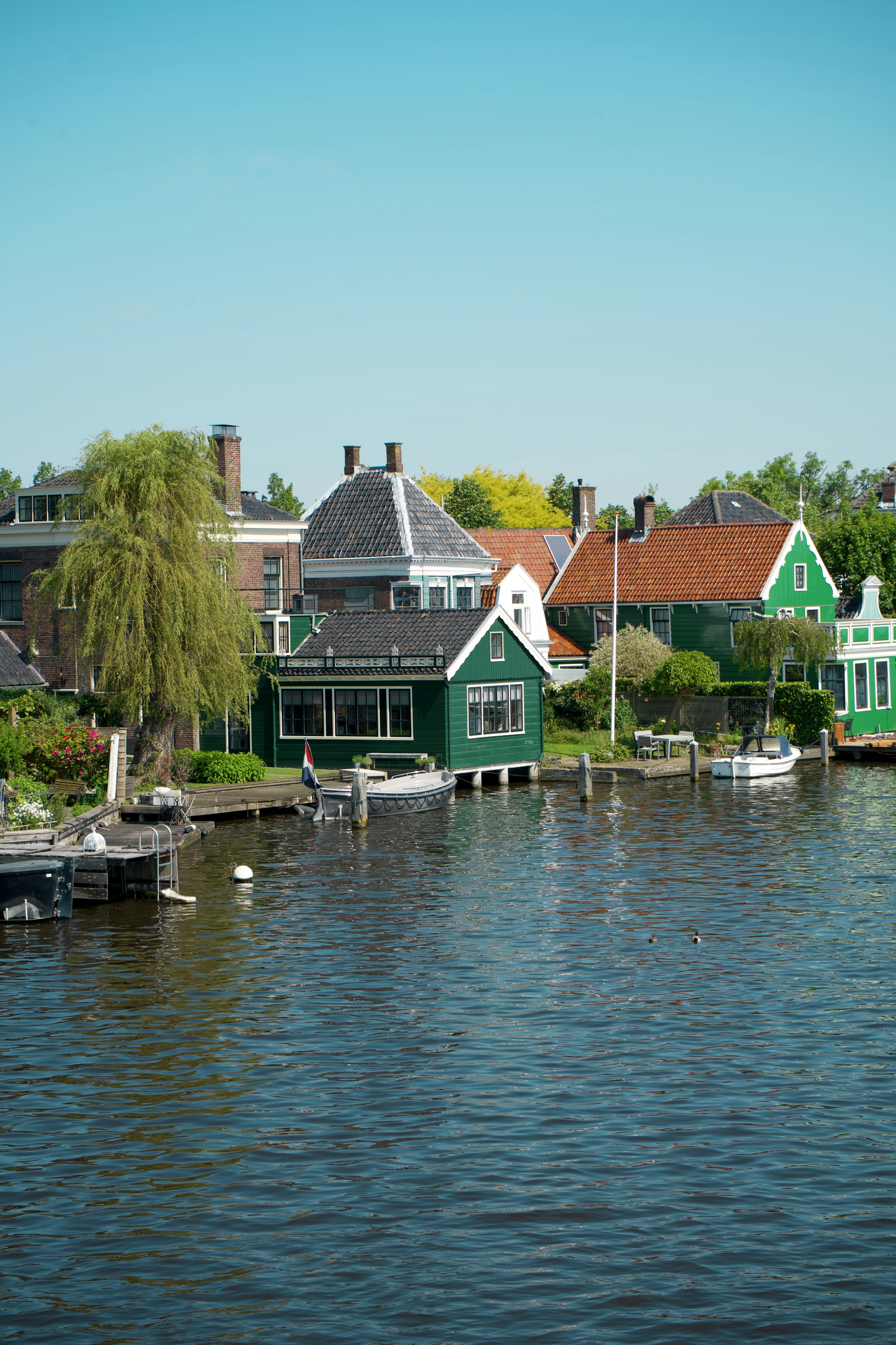 Quaint green and red houses lining a tranquil canal, surrounded by lush greenery and boats gently floating. A perfect representation of Dutch architectural beauty.