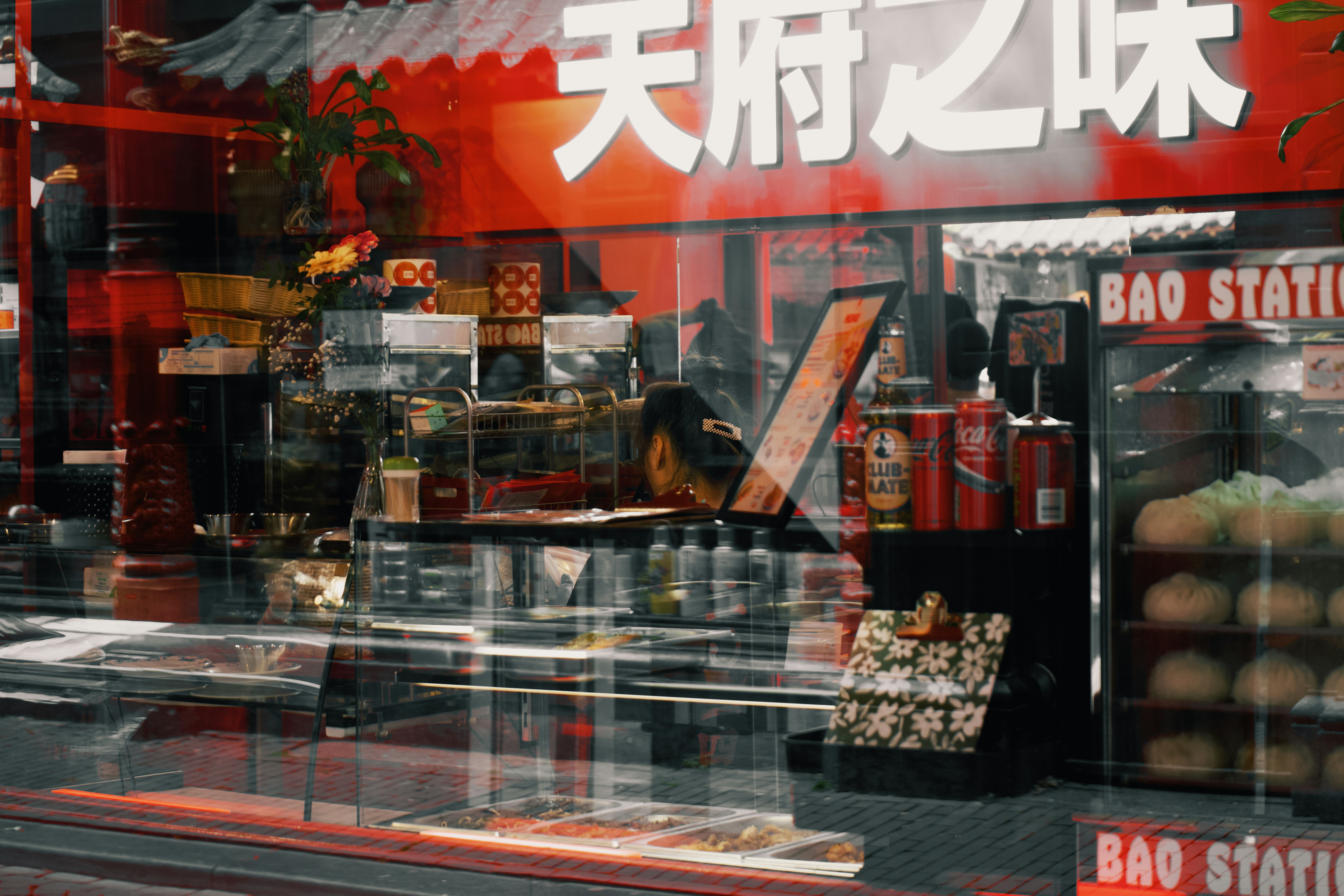 Street reflection in a vibrant bao shop window with red signage and assorted pastries.