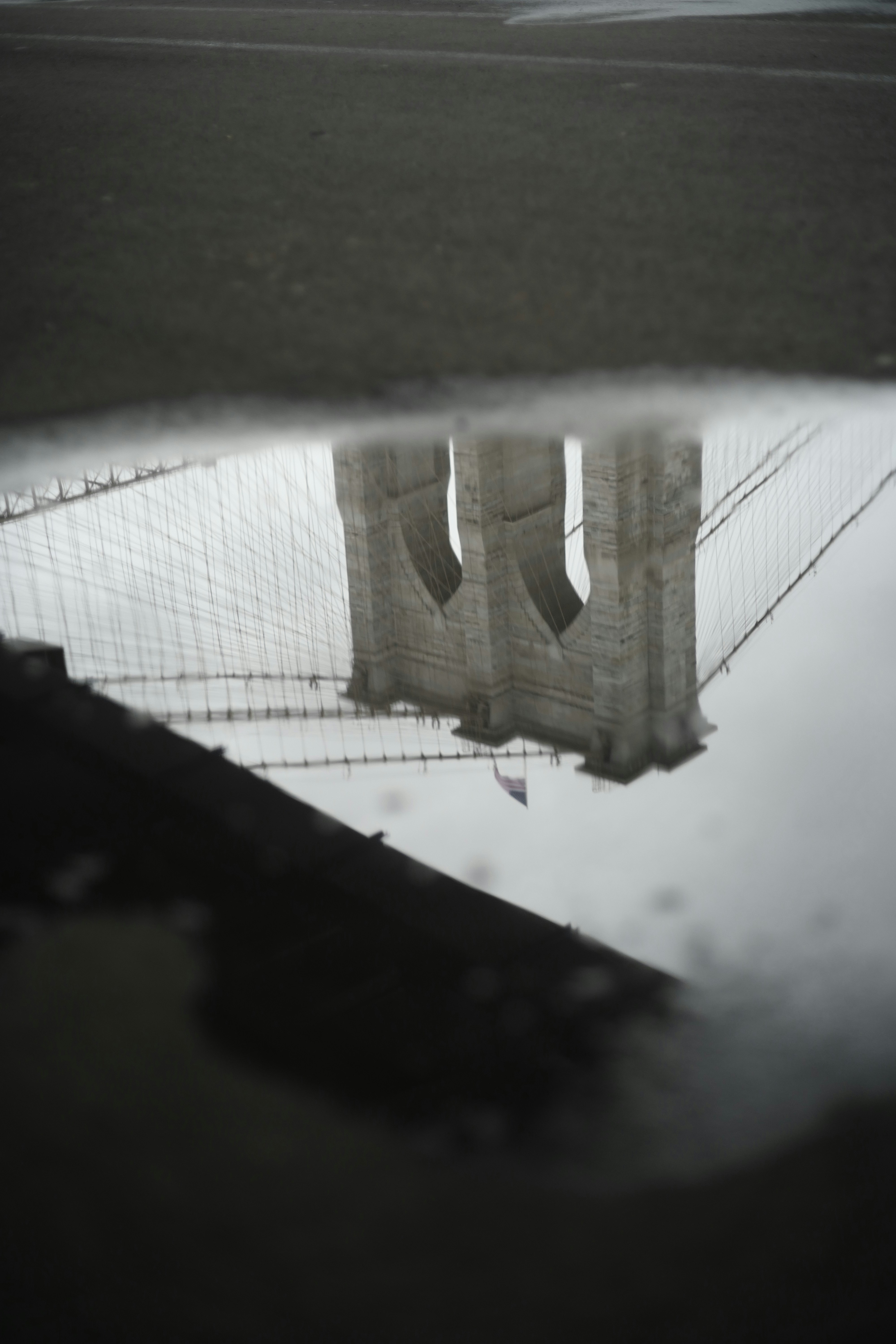 Monochrome photograph capturing a bridge's twin arches reflected in a rain-pooled surface, emphasizing geometric symmetry.