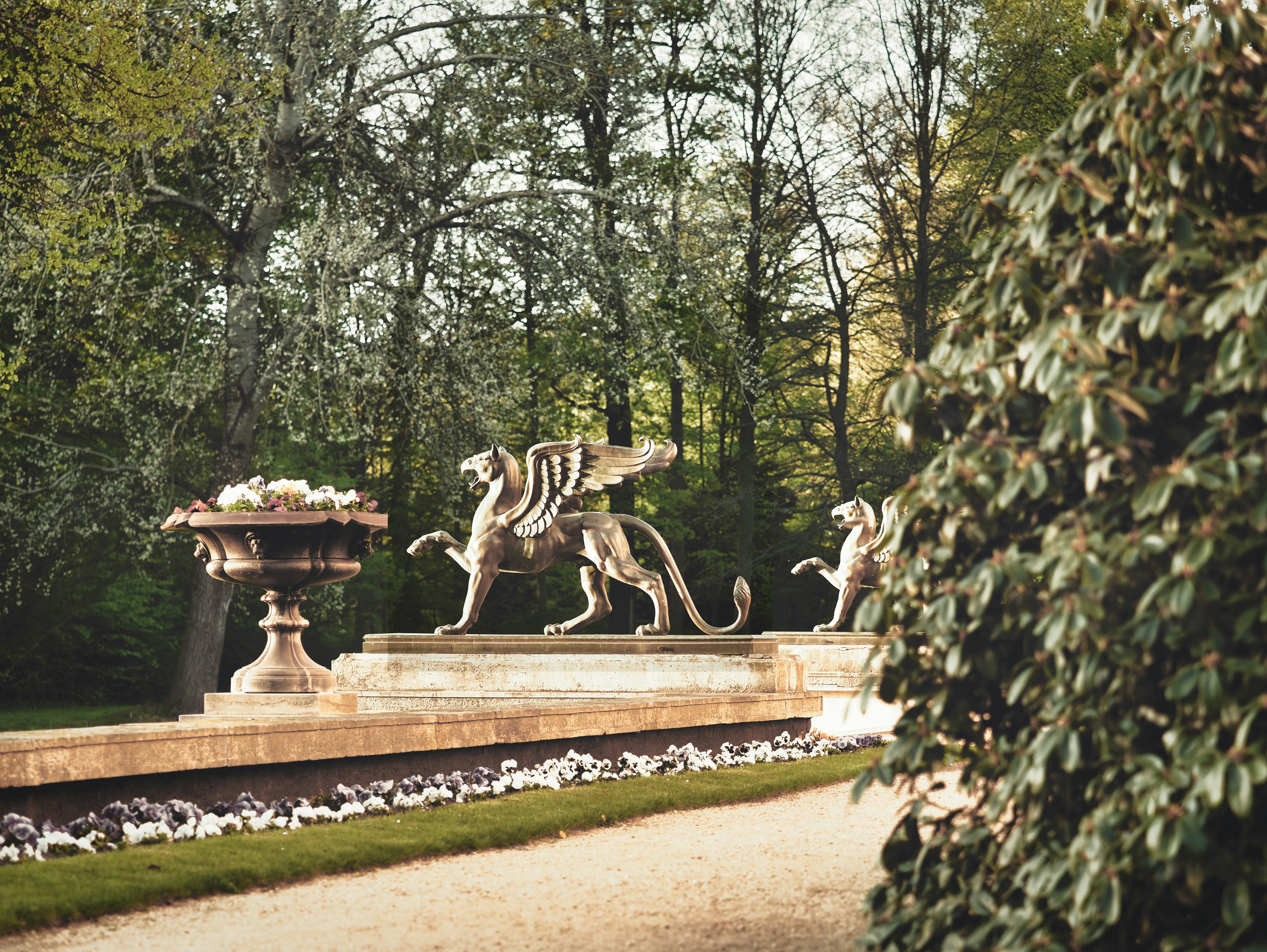 Stone griffin statues stand sentinel along a garden path surrounded by lush greenery.