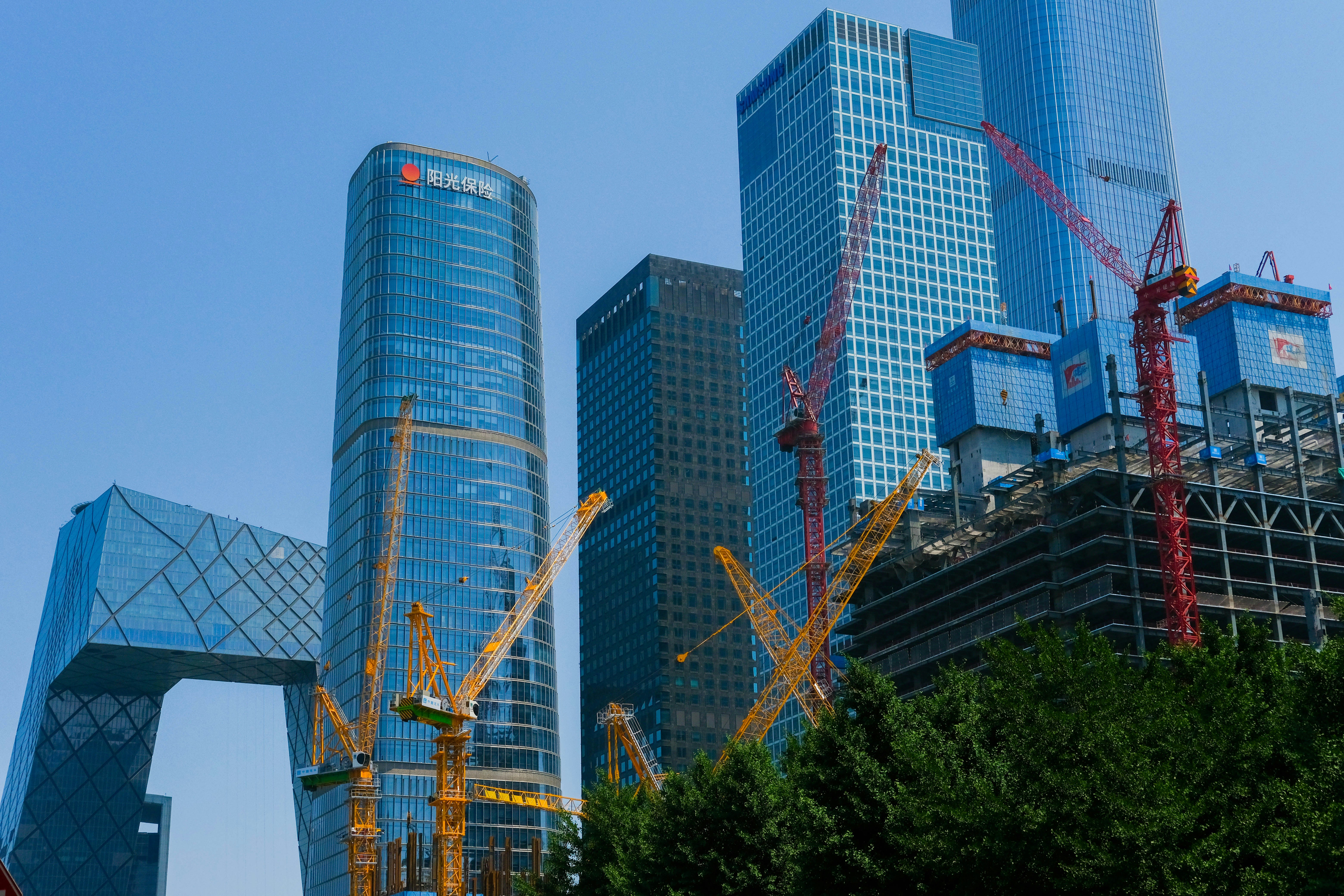 Modern skyscrapers and cranes rise above lush greenery under a clear blue sky.
