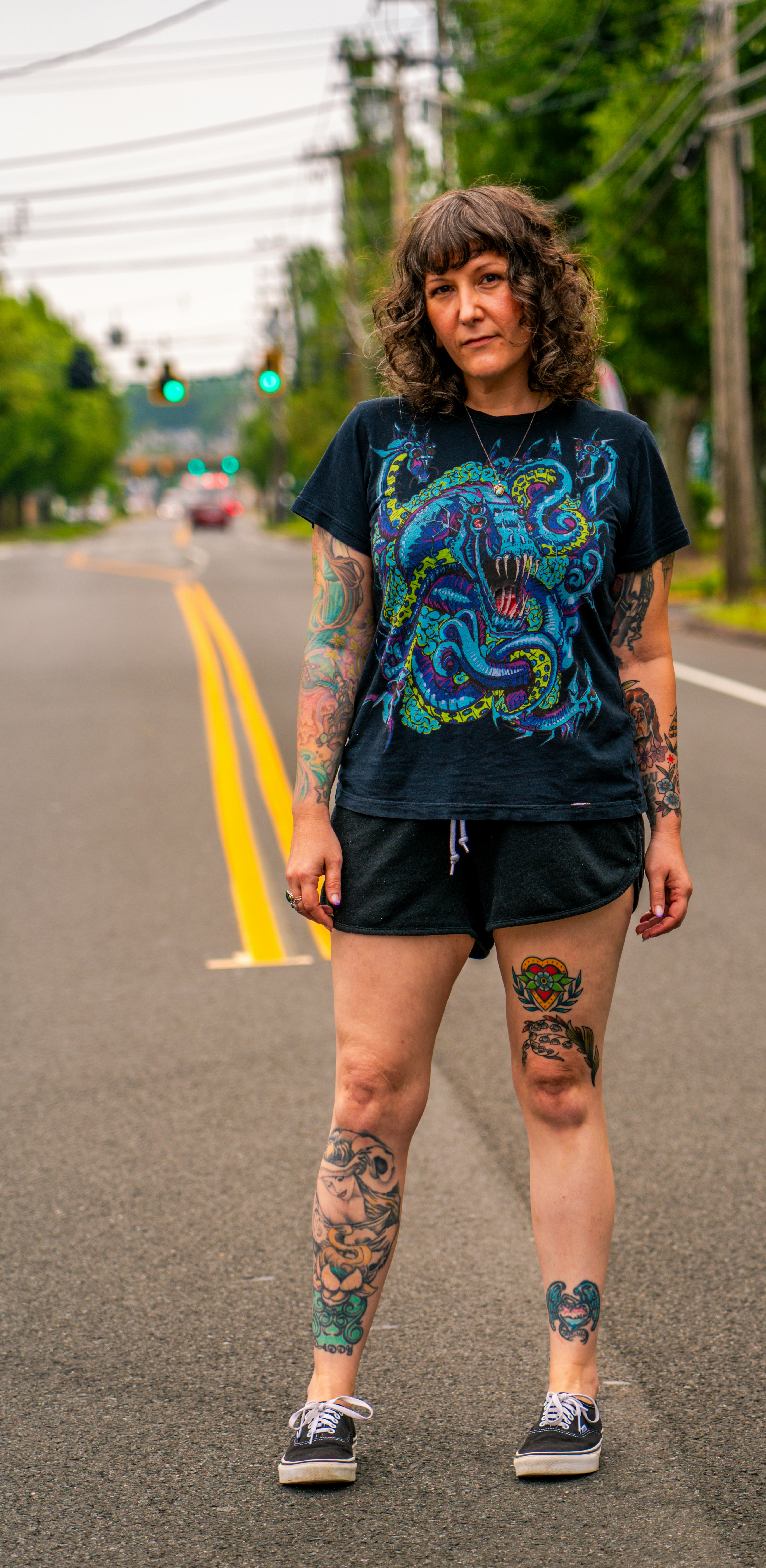 Tattooed woman stands barefoot in the center of a quiet road, wearing a graphic tee and shorts, with a confident gaze as the street fades behind her.