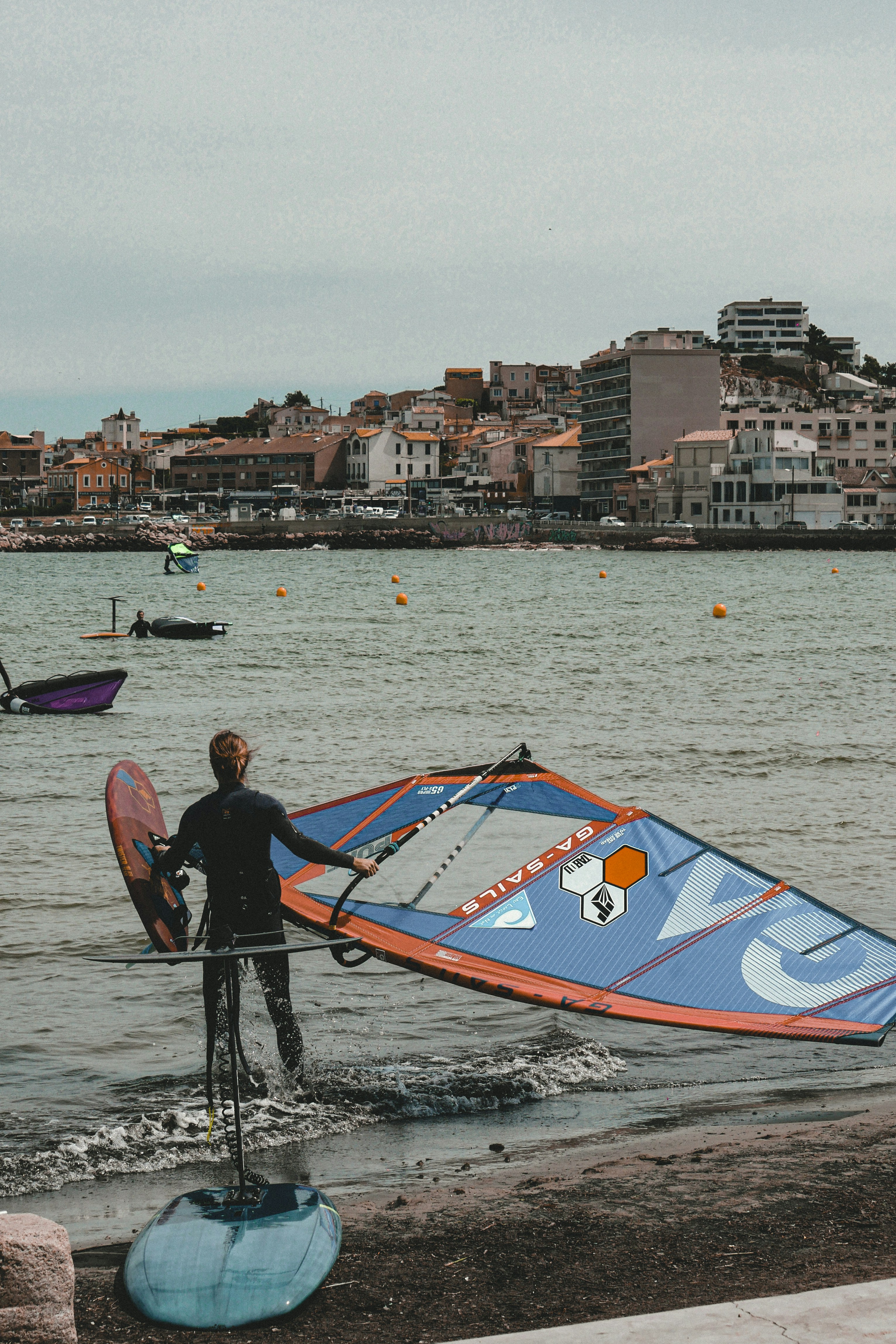Pebble Beach Winds: Windsurfer Aligns a Bright Sail as a Coastal Town Gathers in the Background