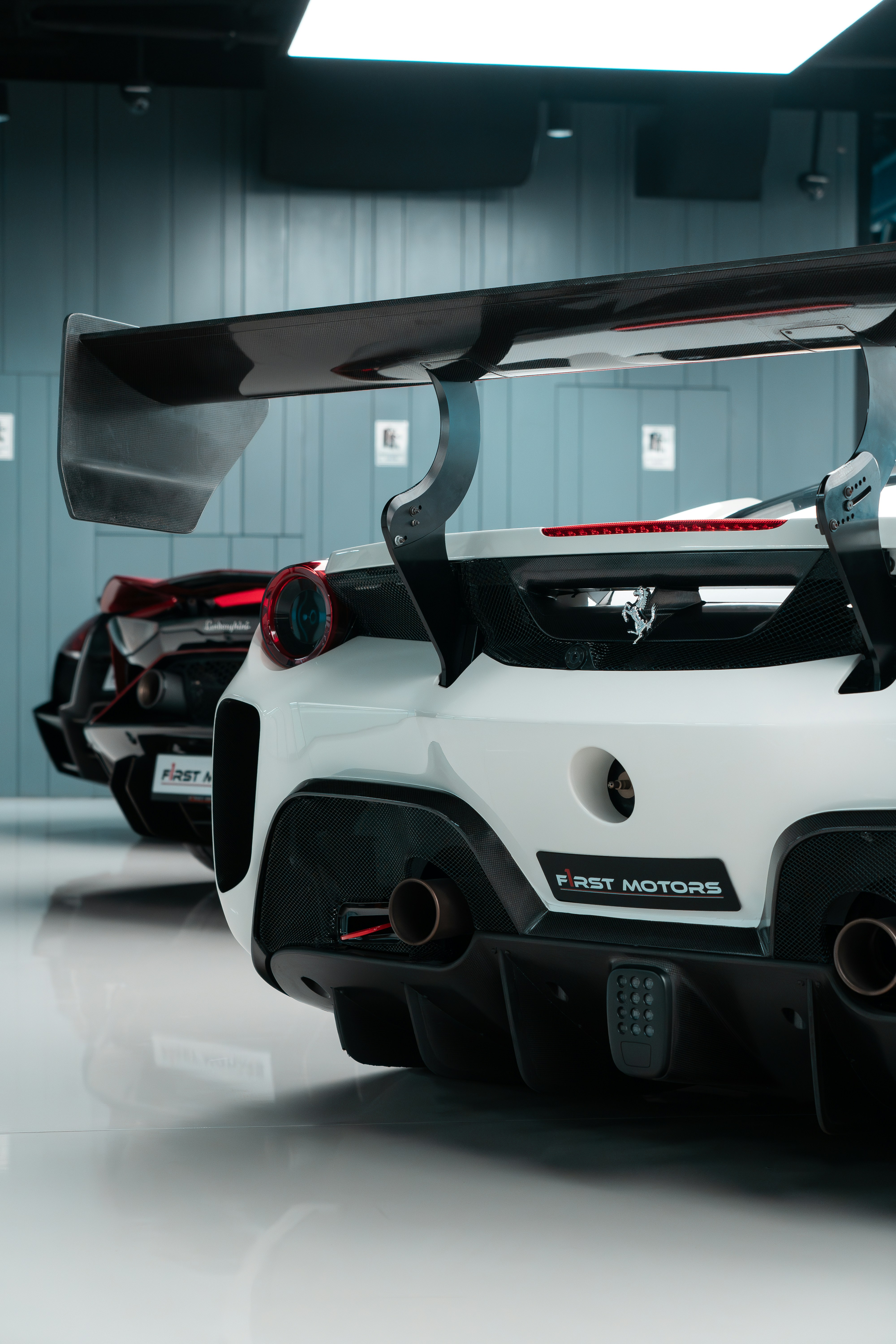 Rear view of a sleek white Ferrari with a prominent spoiler, showcasing its aerodynamic features alongside a dark-colored Ferrari in the background. 