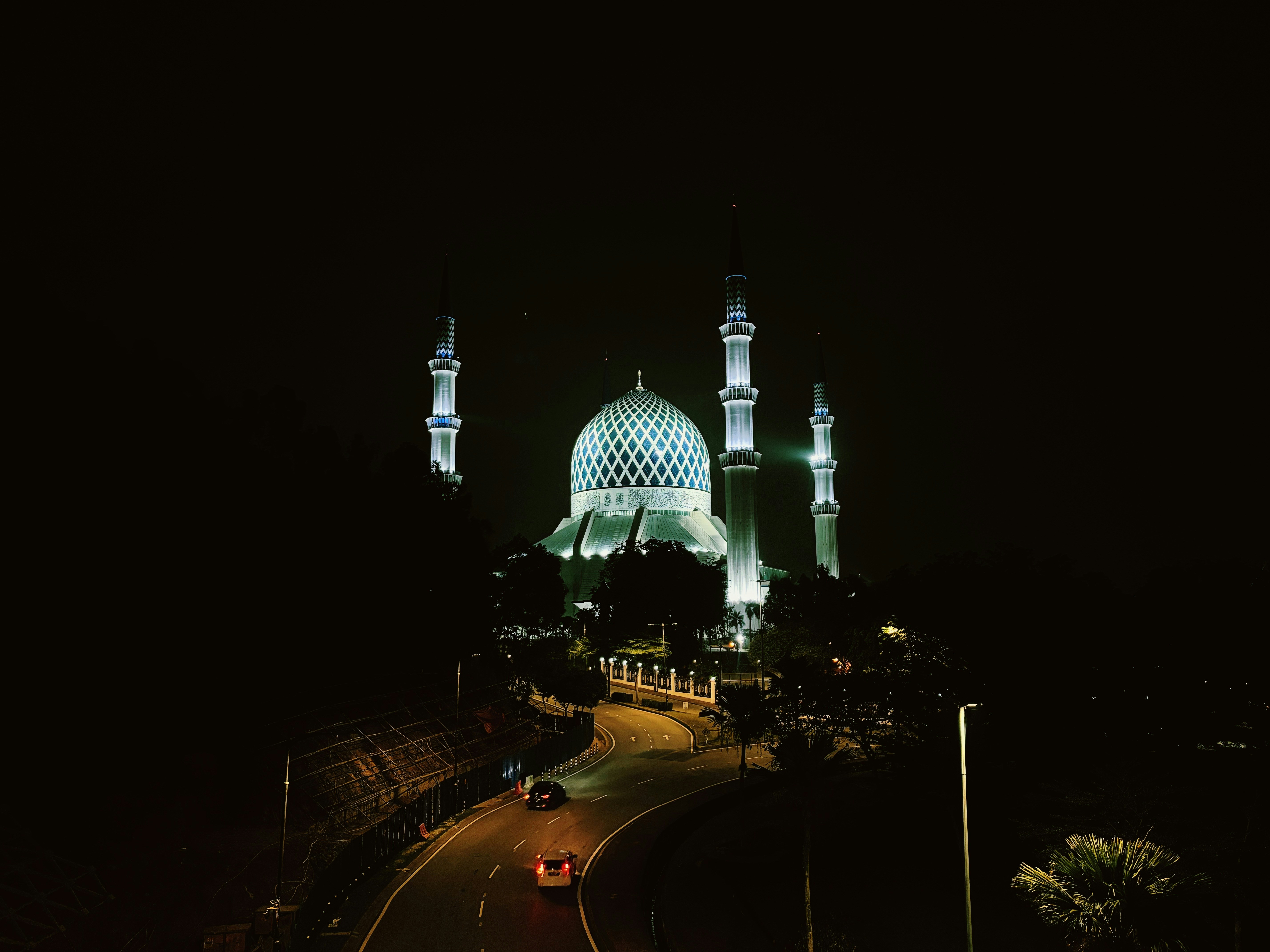 A grand mosque with glowing domes and minarets stands illuminated against the night sky, with a winding road leading towards it.